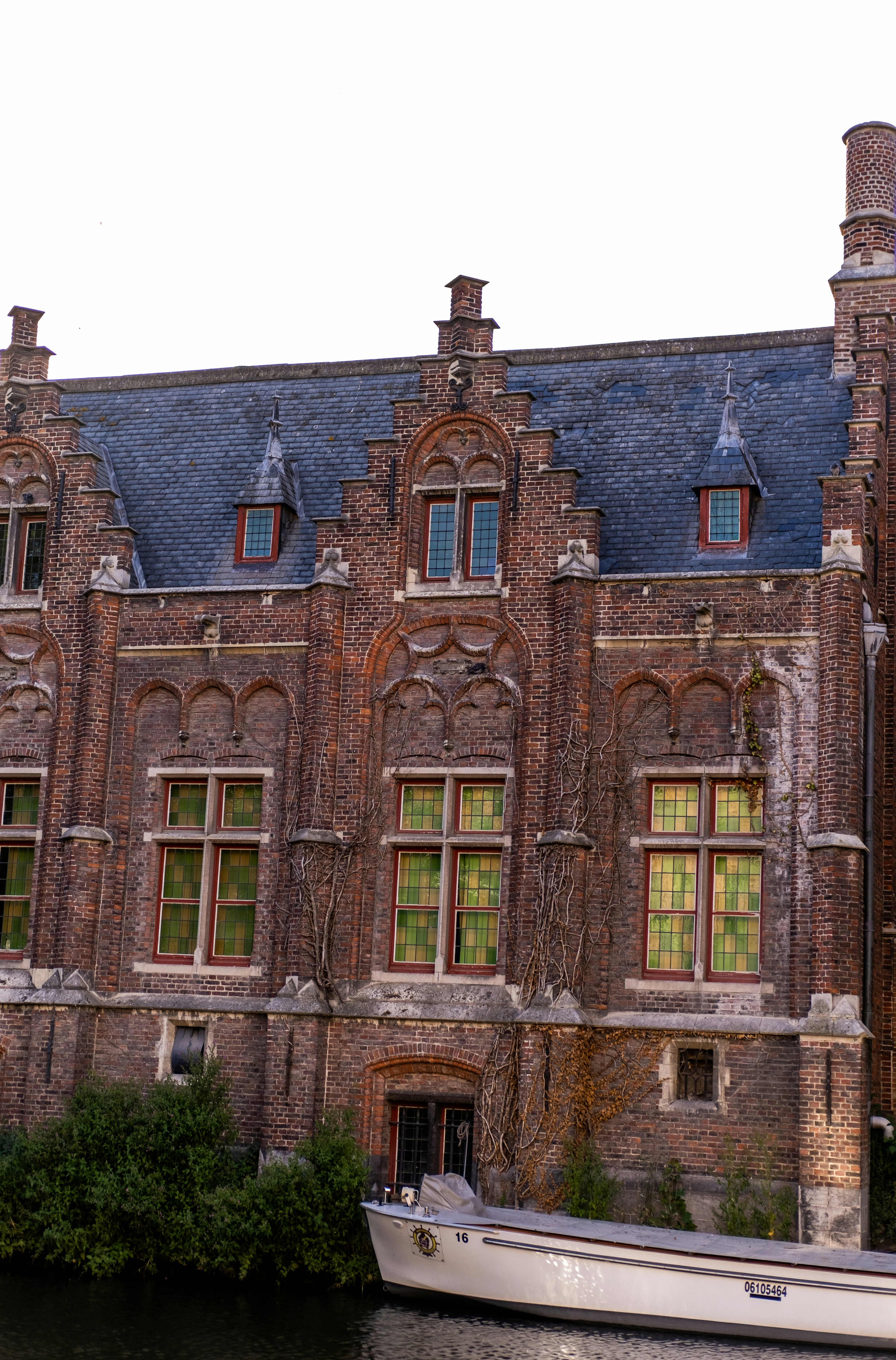 Historic brick building adorned with colorful windows, partially veiled by creeping vines, alongside a serene waterway with a boat moored nearby.