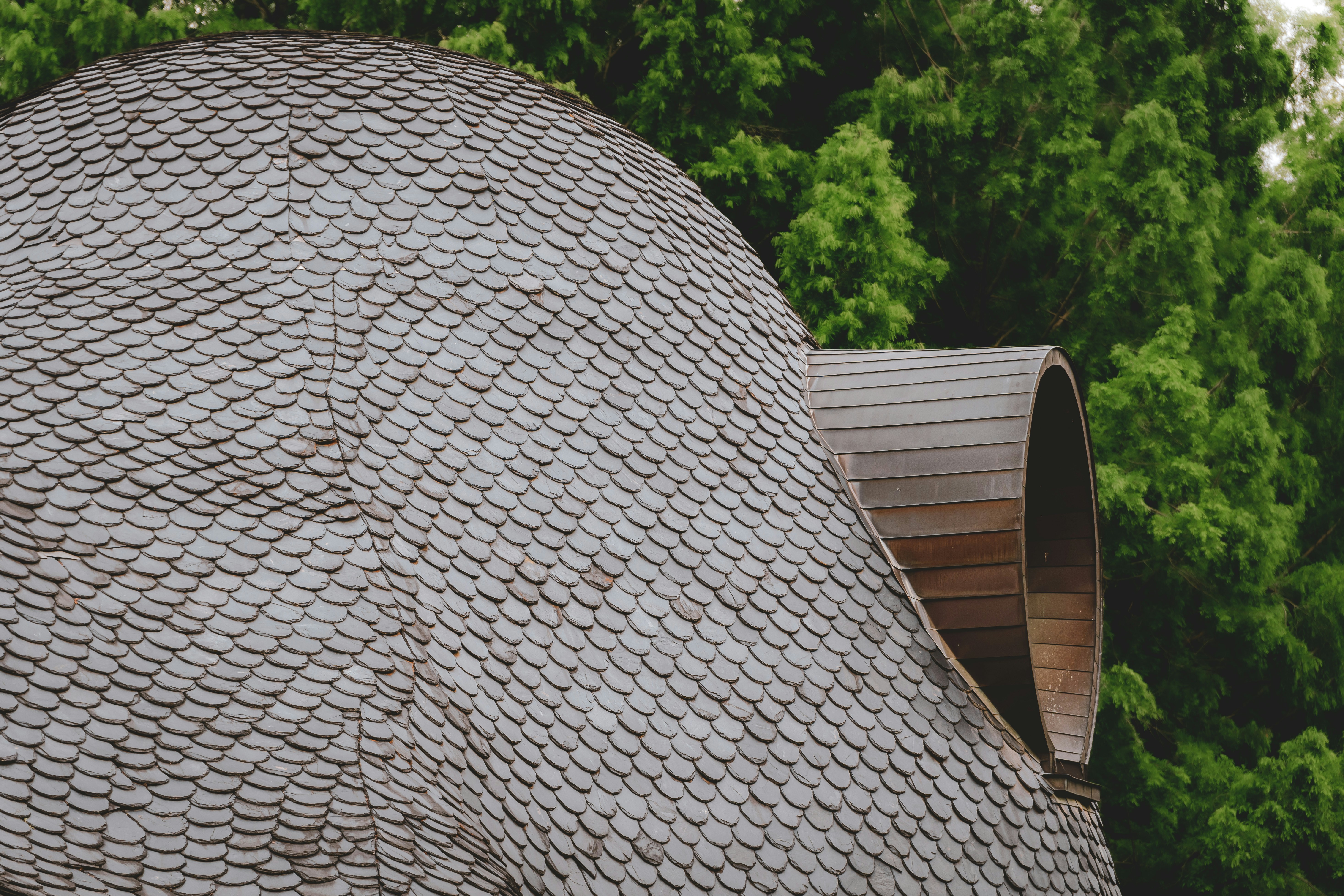 Textured domed roof covered in overlapping grey shingles with a cylindrical vent on the right, set against a backdrop of green trees.