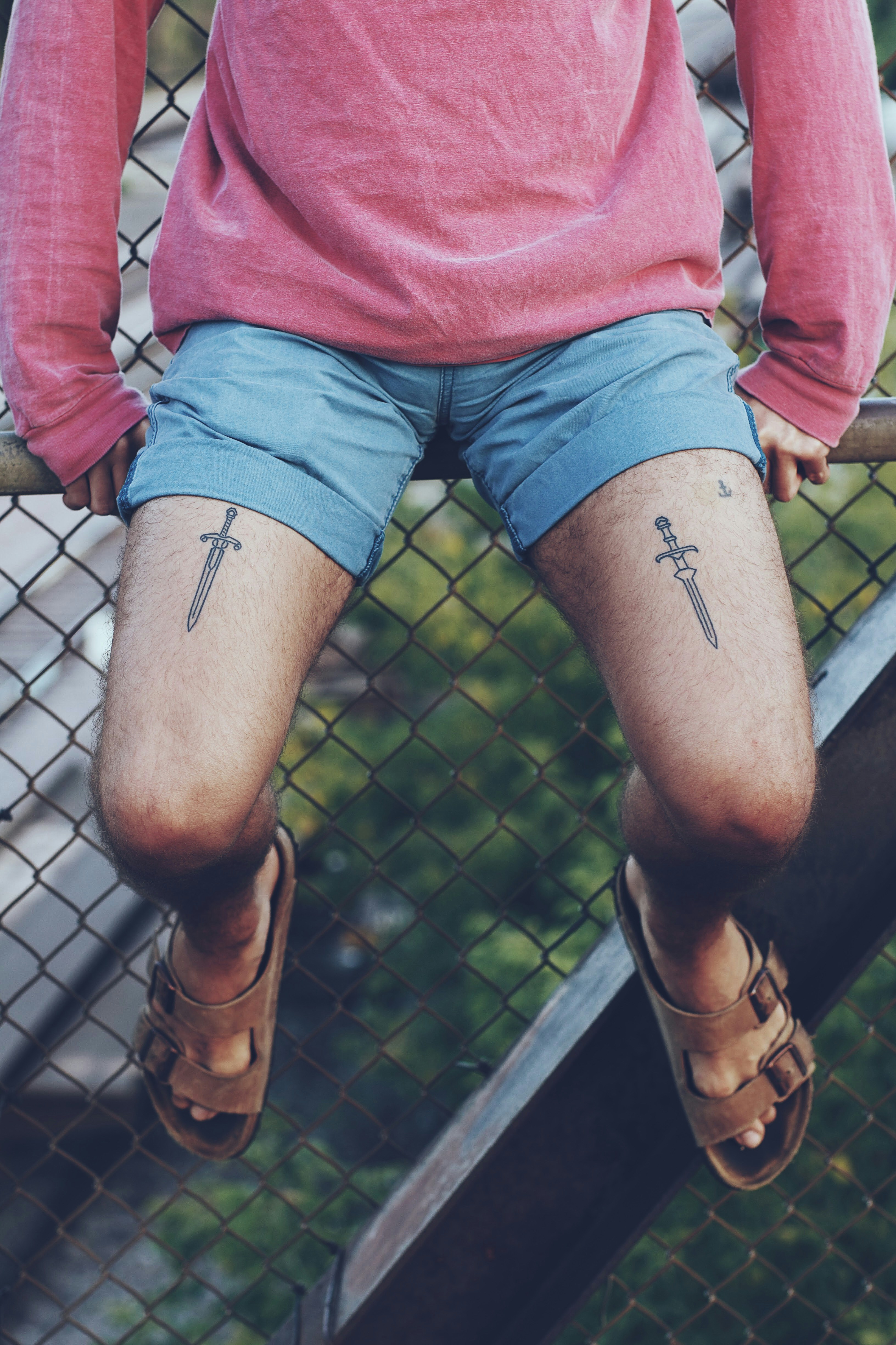 Twin Swords on Sunlit Thighs Behind a Chain-Link Fence
