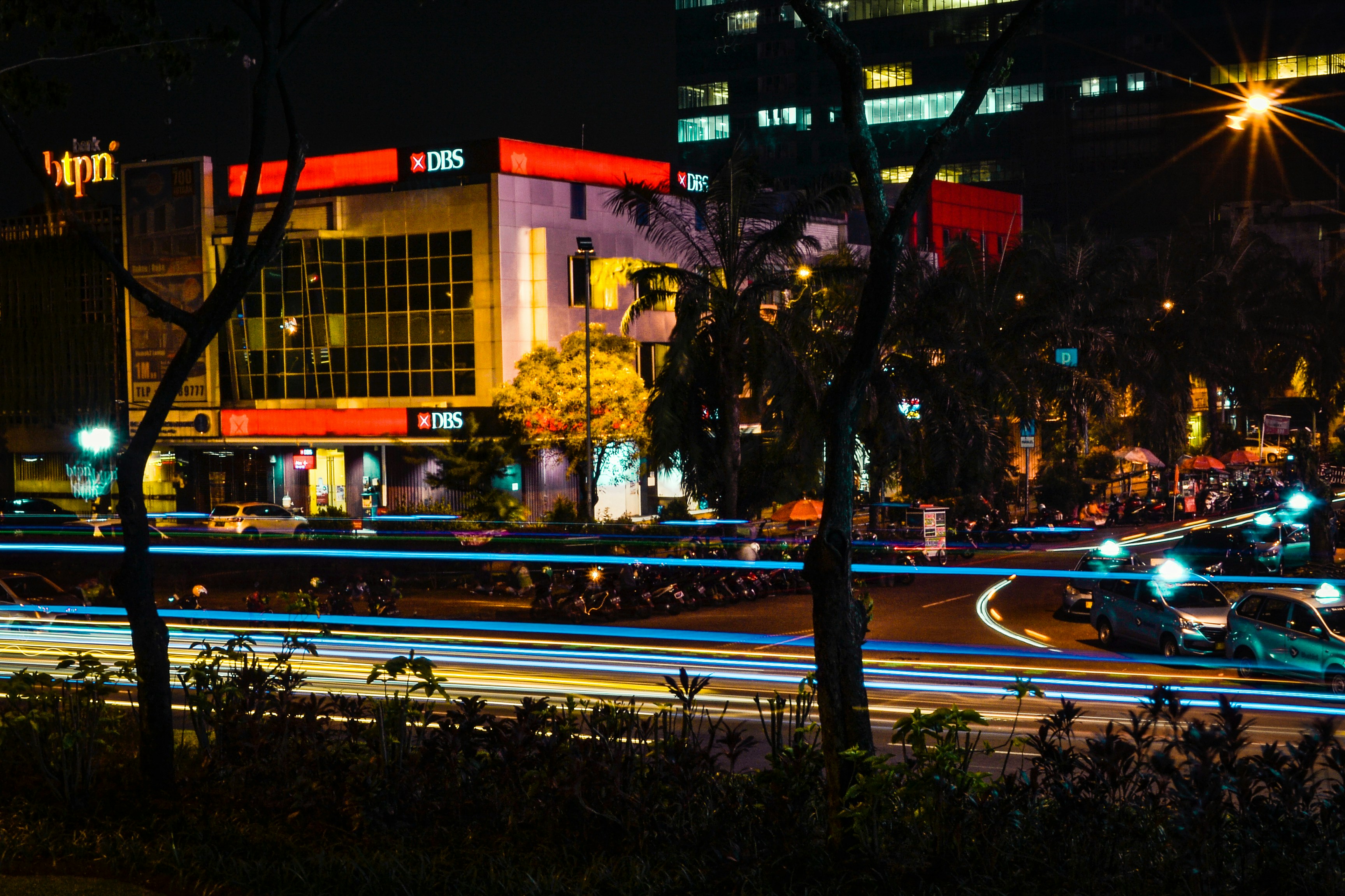 Nighttime city street with light trails from passing vehicles, framed by silhouettes of trees and illuminated buildings in the background.