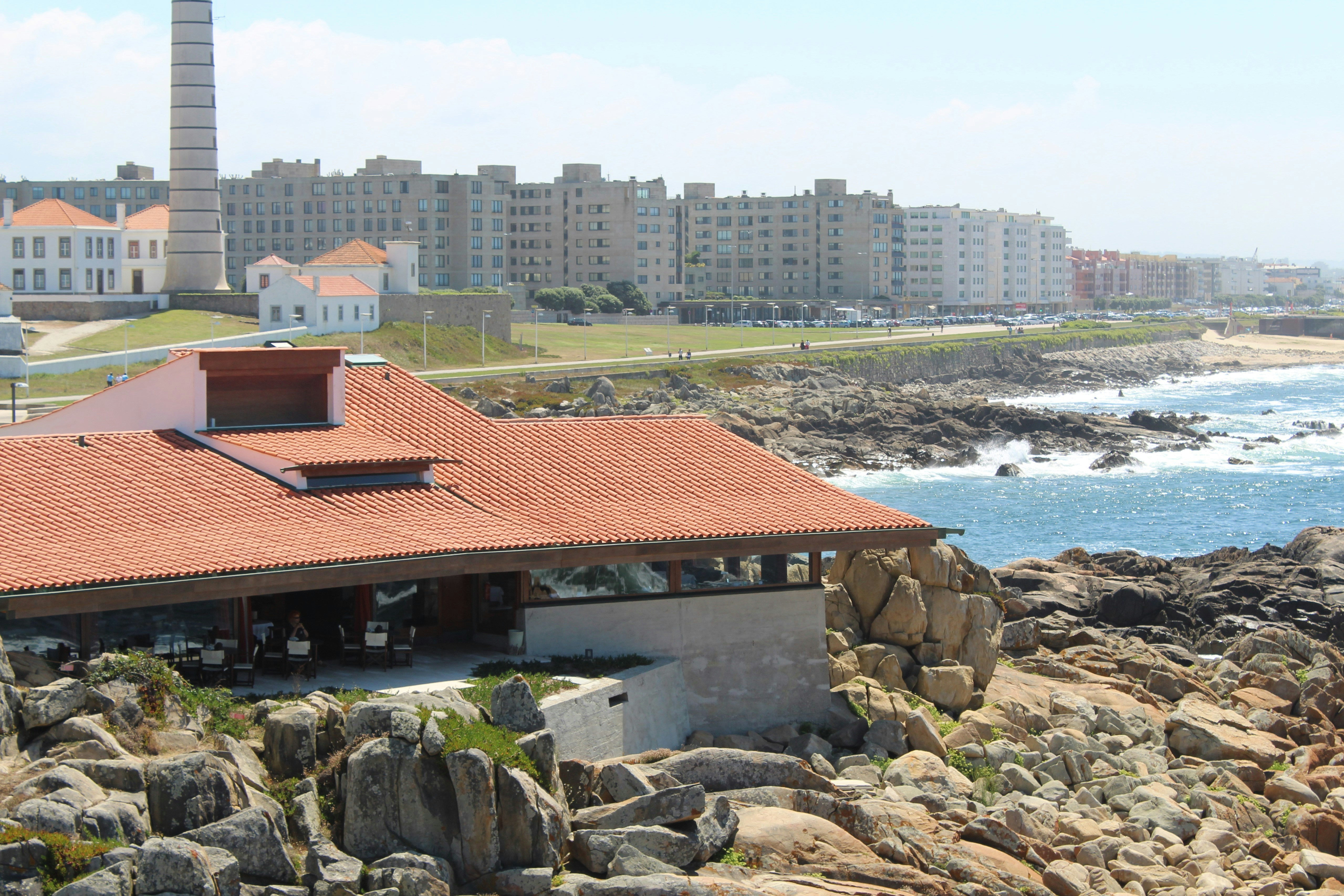 Modern building with a red roof nestled among rocky coastal terrain, with urban skyline in the background.