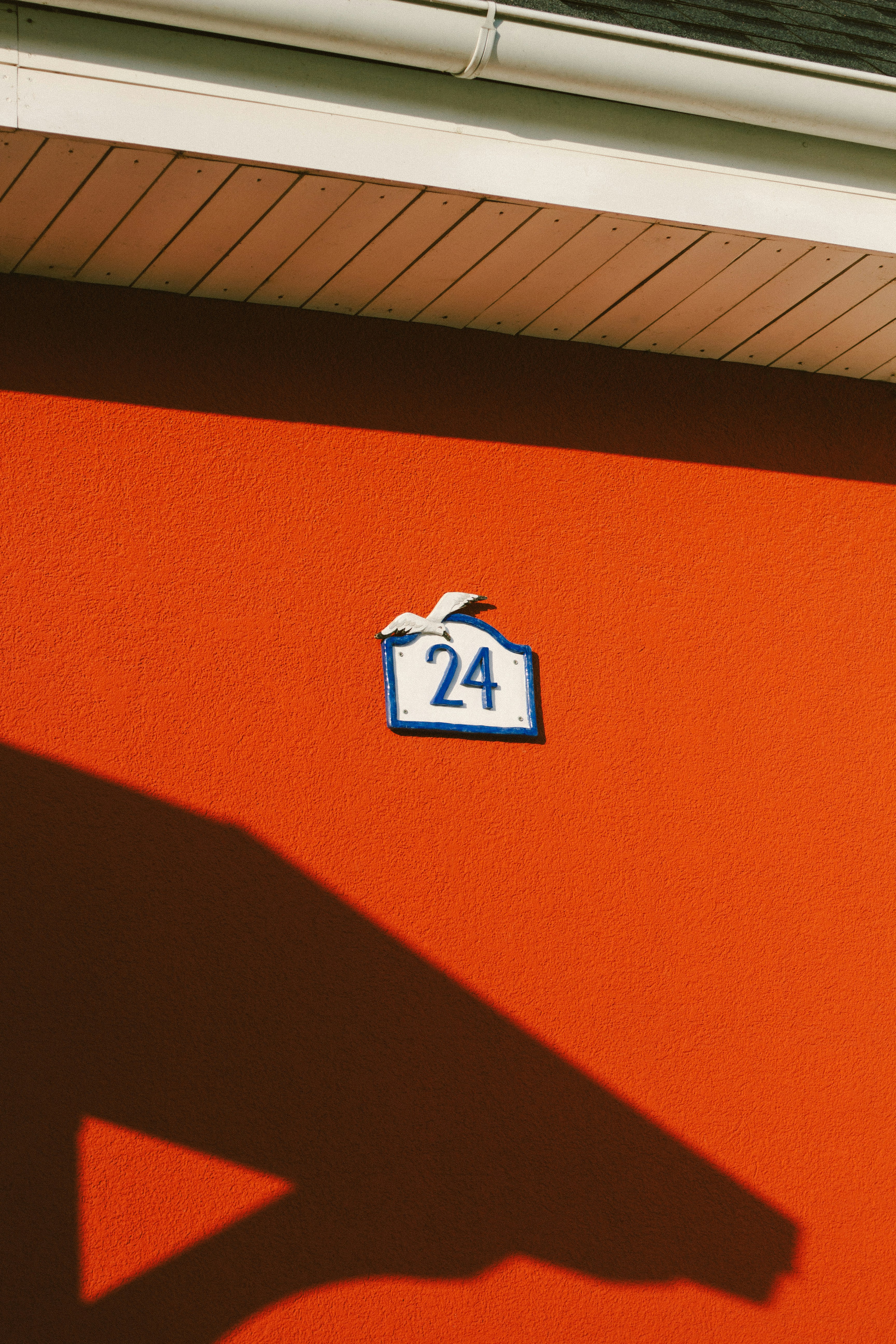 House number 24 displayed on a bright orange wall, with a subtle bird motif above the digits. The shadows cast create an interesting interplay of shapes.