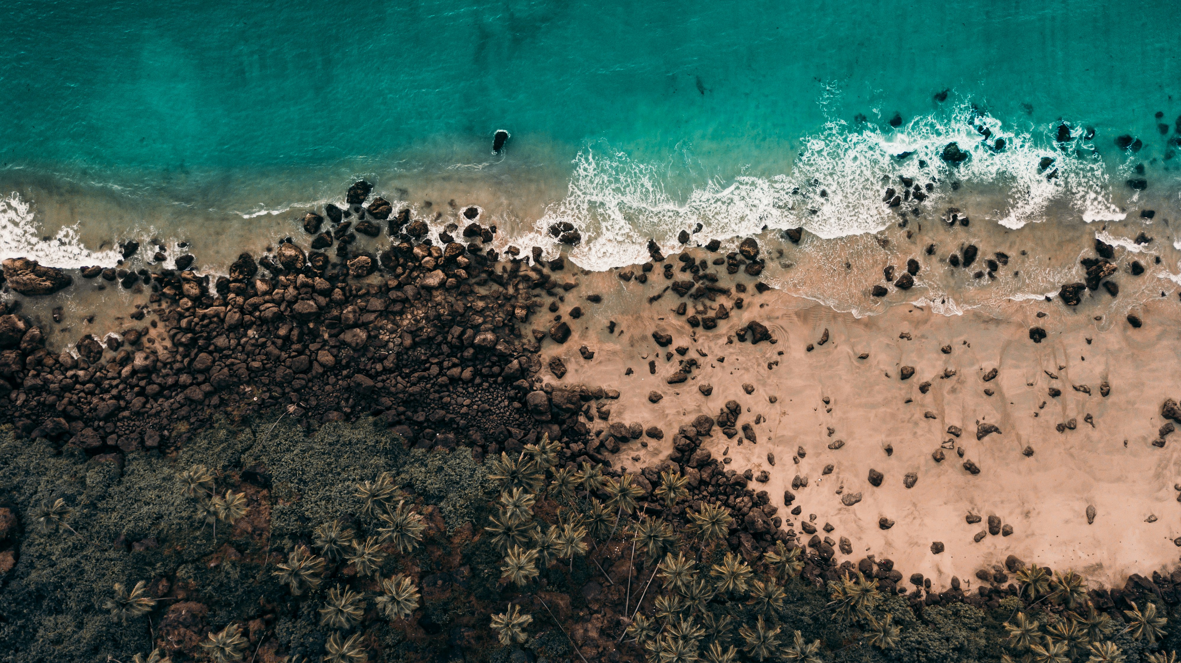 Aerial view of a rocky coastline meeting a tranquil beach, surrounded by lush greenery and gentle waves lapping at the shore.