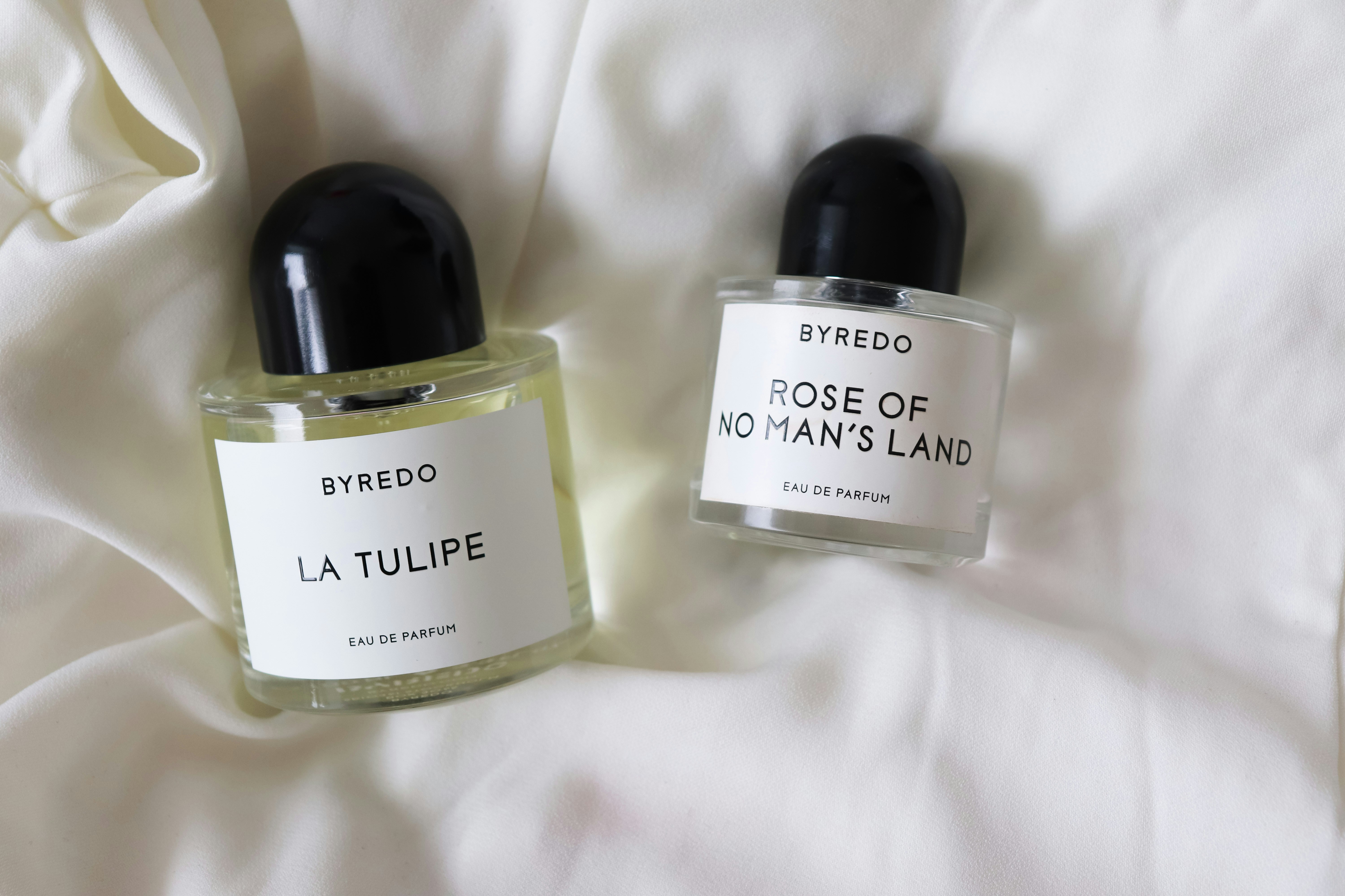 Two Byredo perfume bottles rest on white fabric, soft shadows and gentle lighting emphasize label typography and glass contours.
