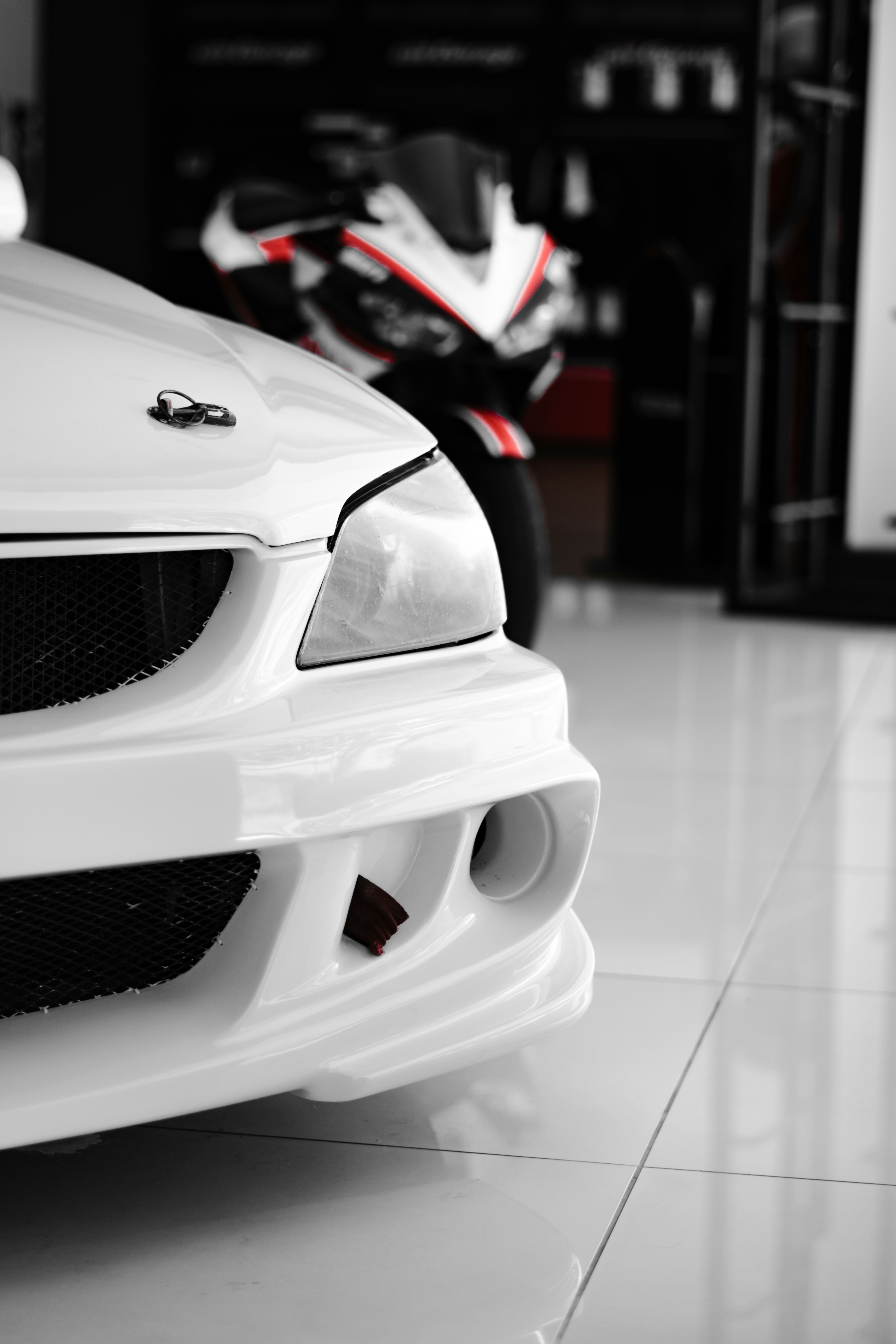 Close-up of a sleek white car's front end, showcasing its design against a blurred motorcycle backdrop. The contrast highlights the car's aggressive styling.