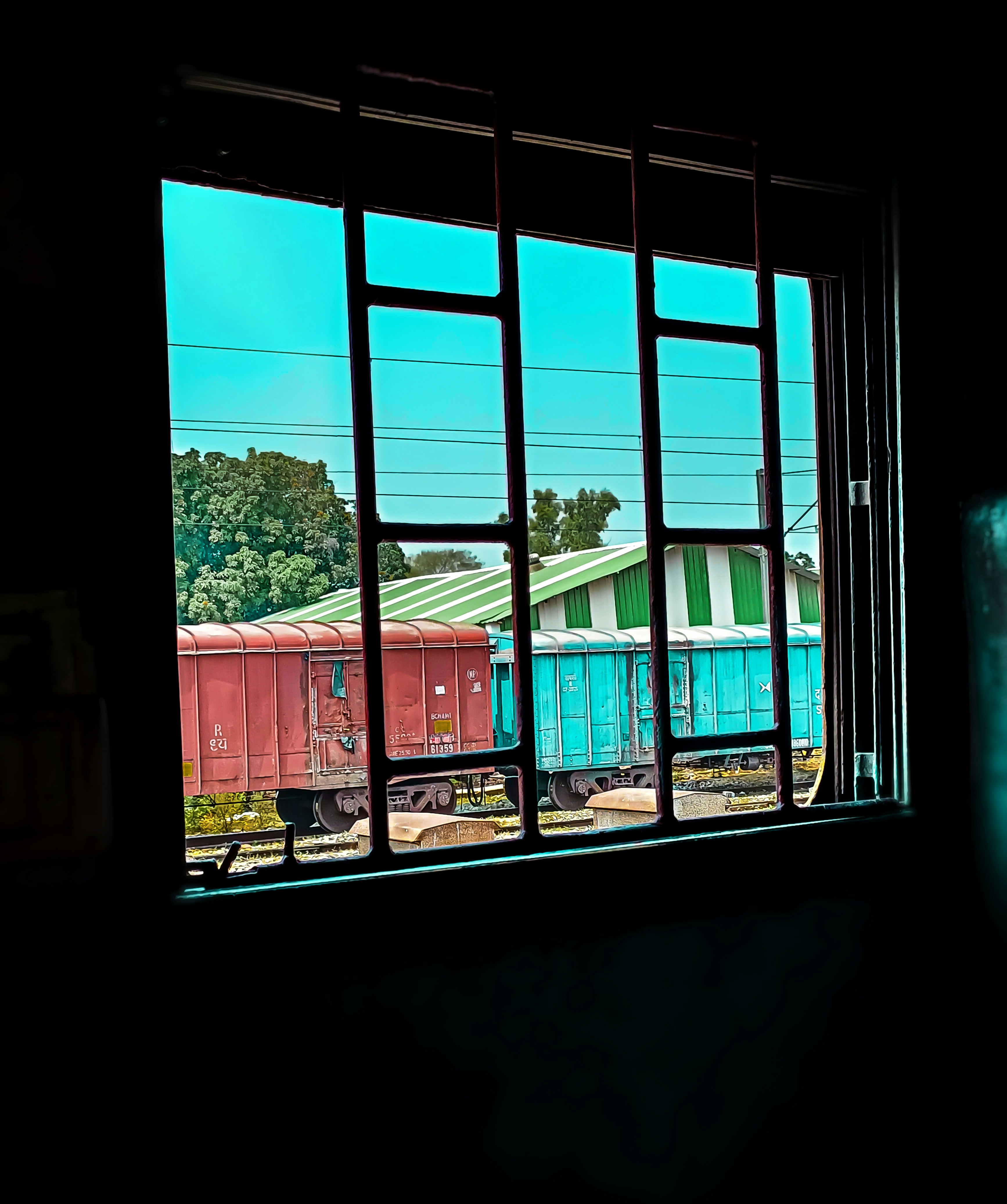 View through a barred window showcasing colorful freight trains against a backdrop of lush trees and a green-roofed building.