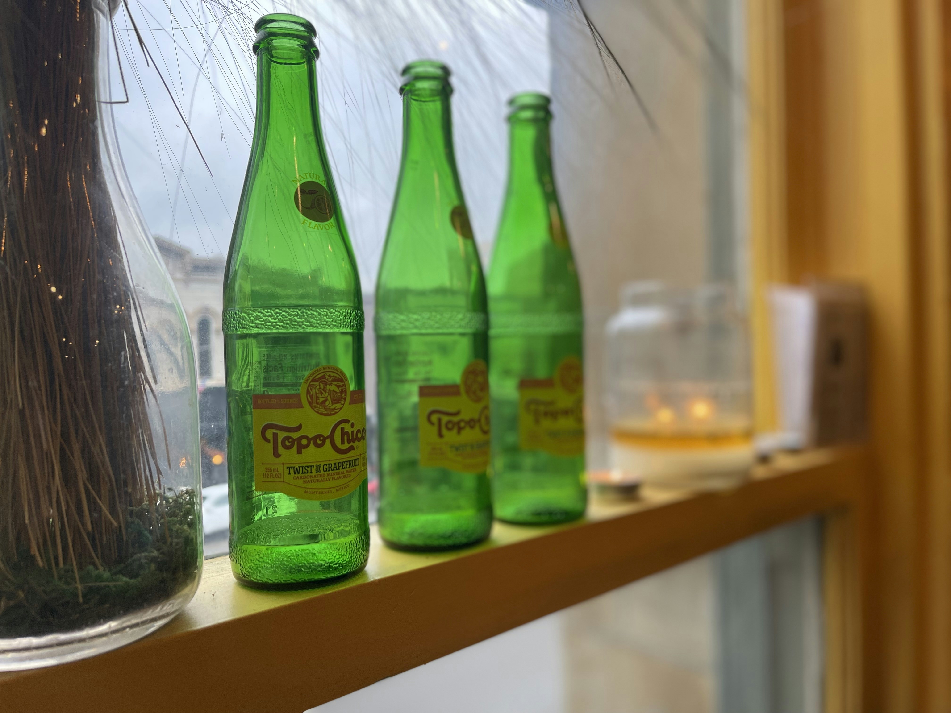 Three green Topo Chico bottles arranged on a windowsill, illuminated by soft natural light. A decorative glass jar and a hint of foliage add depth to the scene.