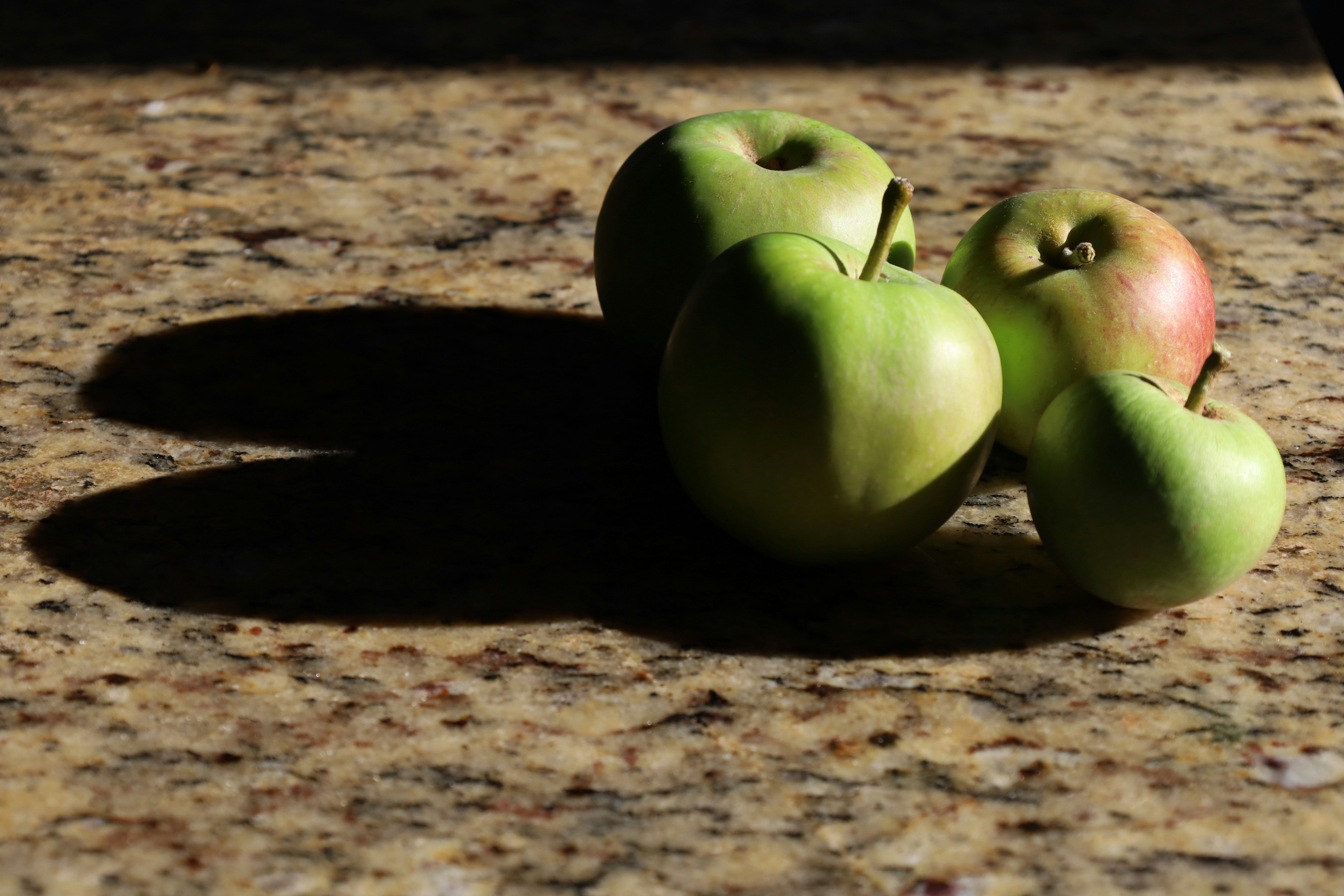 Four green apples resting on a textured granite surface, casting long shadows in soft light.