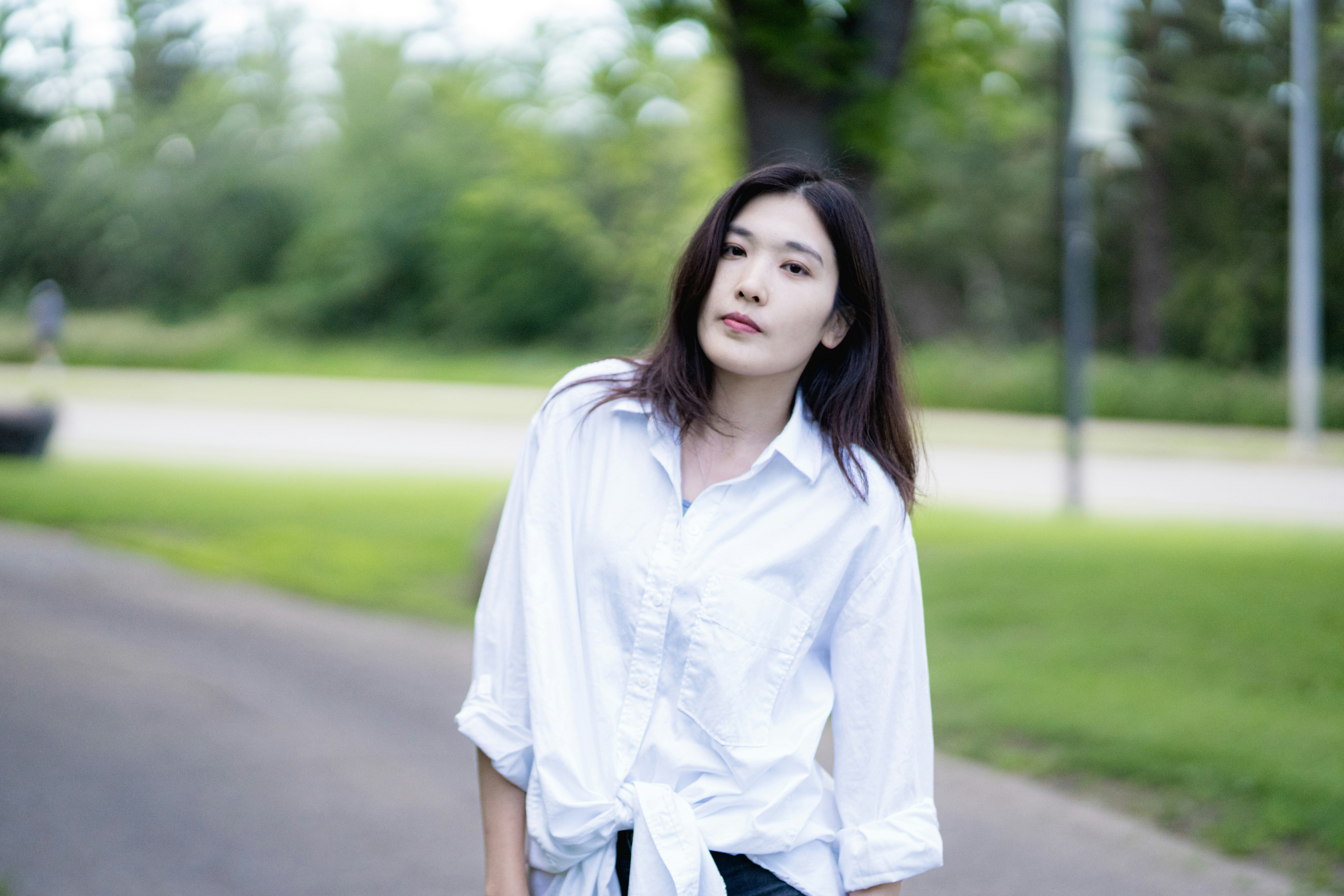 A young woman in a white shirt stands casually on a pathway, surrounded by lush greenery. The soft focus and natural light create a tranquil atmosphere.