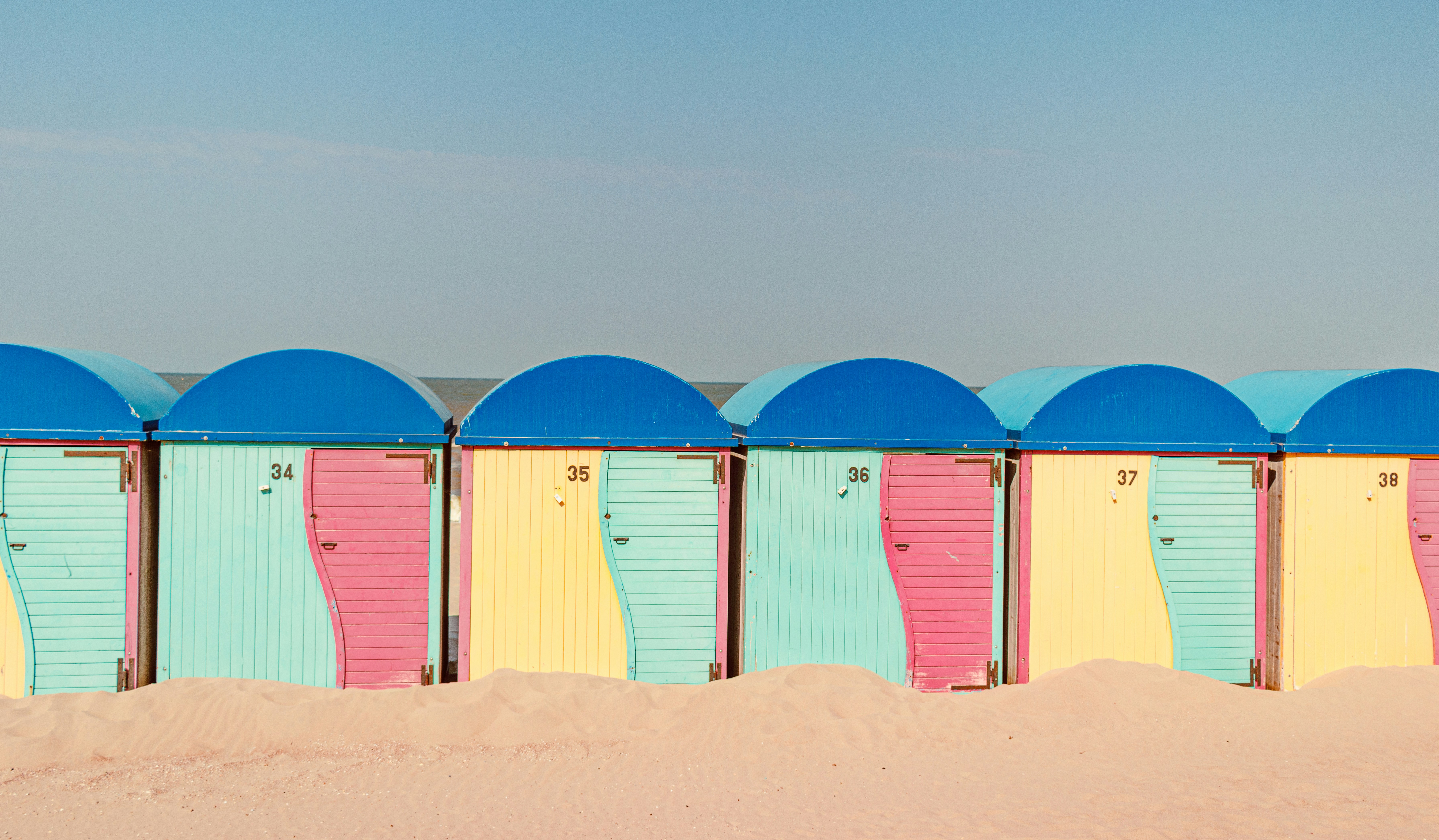 Vibrant beach huts lined along a sandy shore, showcasing a palette of pastel colors and curved roofs. The scene captures the essence of seaside leisure.