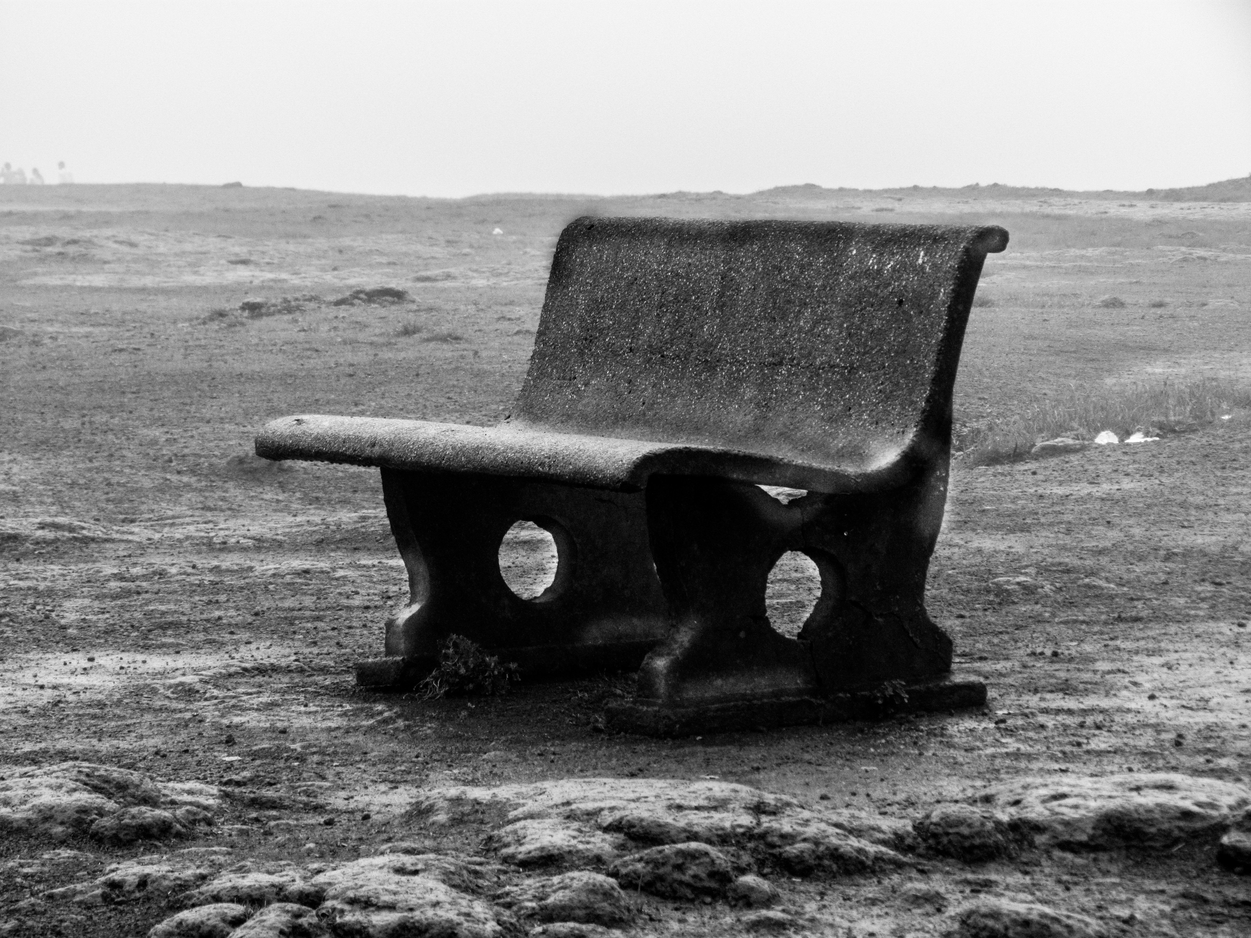 Monochrome photograph of a weathered bench on a wind-swept, barren plain beneath a pale horizon.