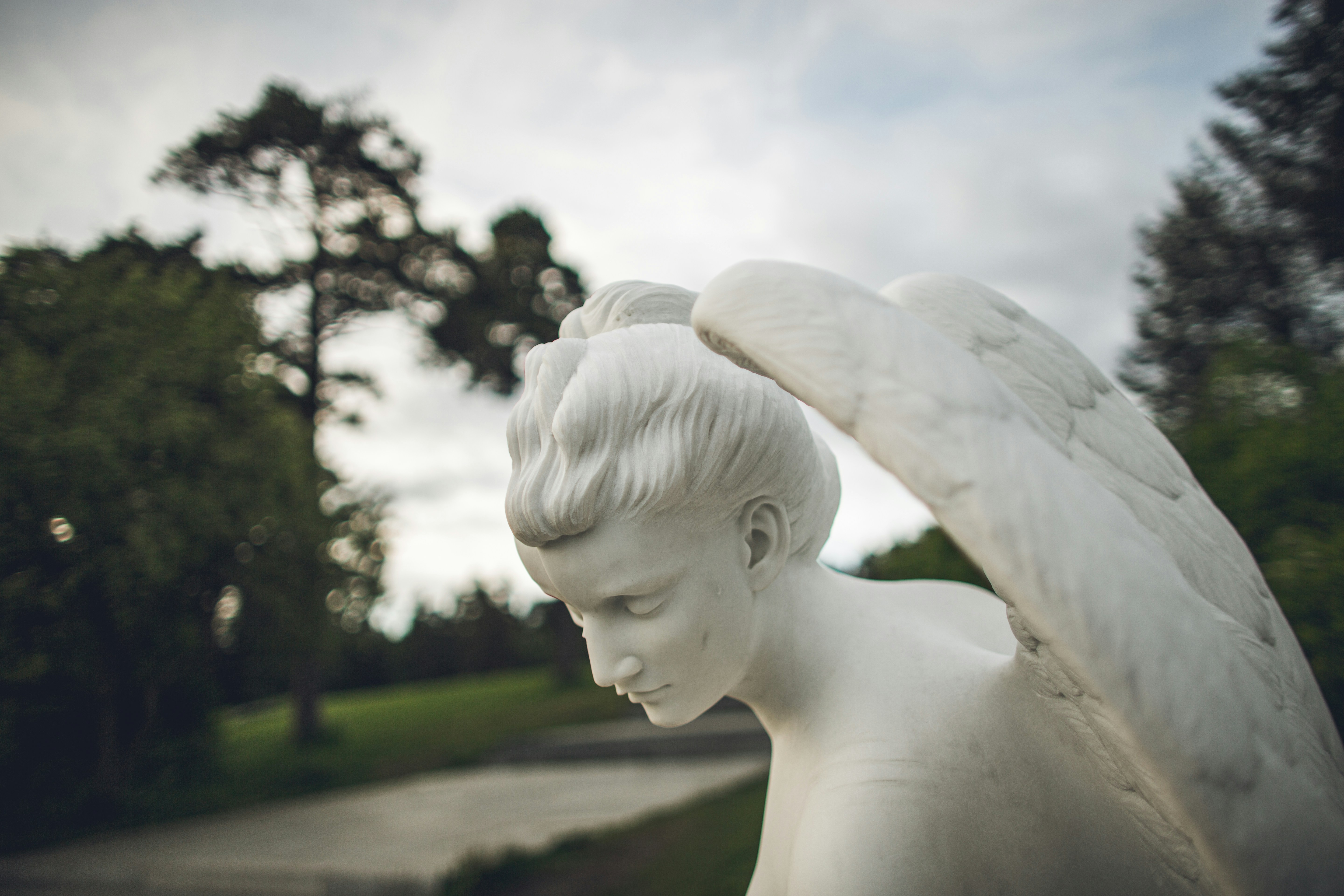 Marble statue of a winged figure in a tranquil park setting under a cloudy sky.