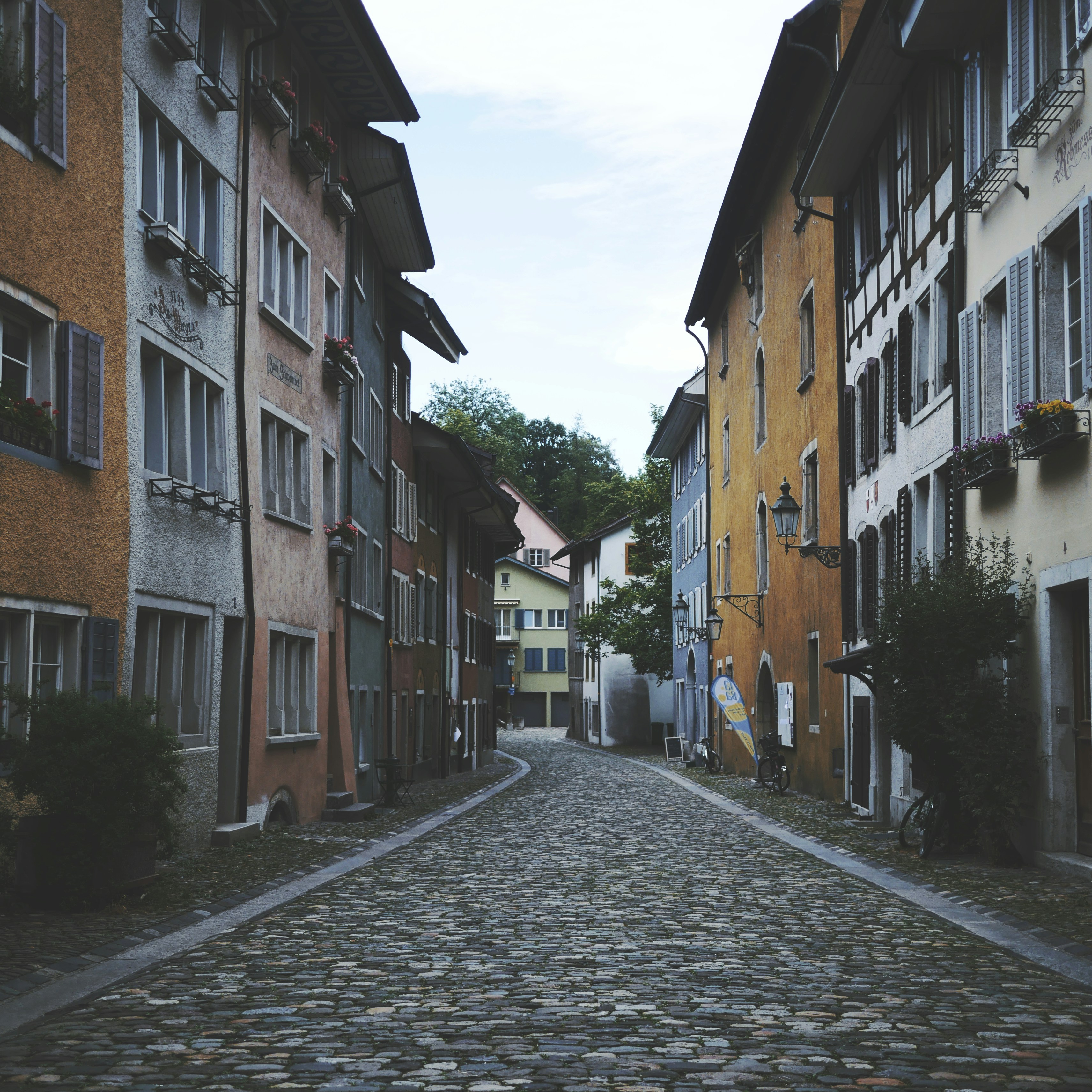 Charming cobblestone street flanked by colorful buildings, leading to a hidden courtyard. Lush greenery peeks through the architecture.