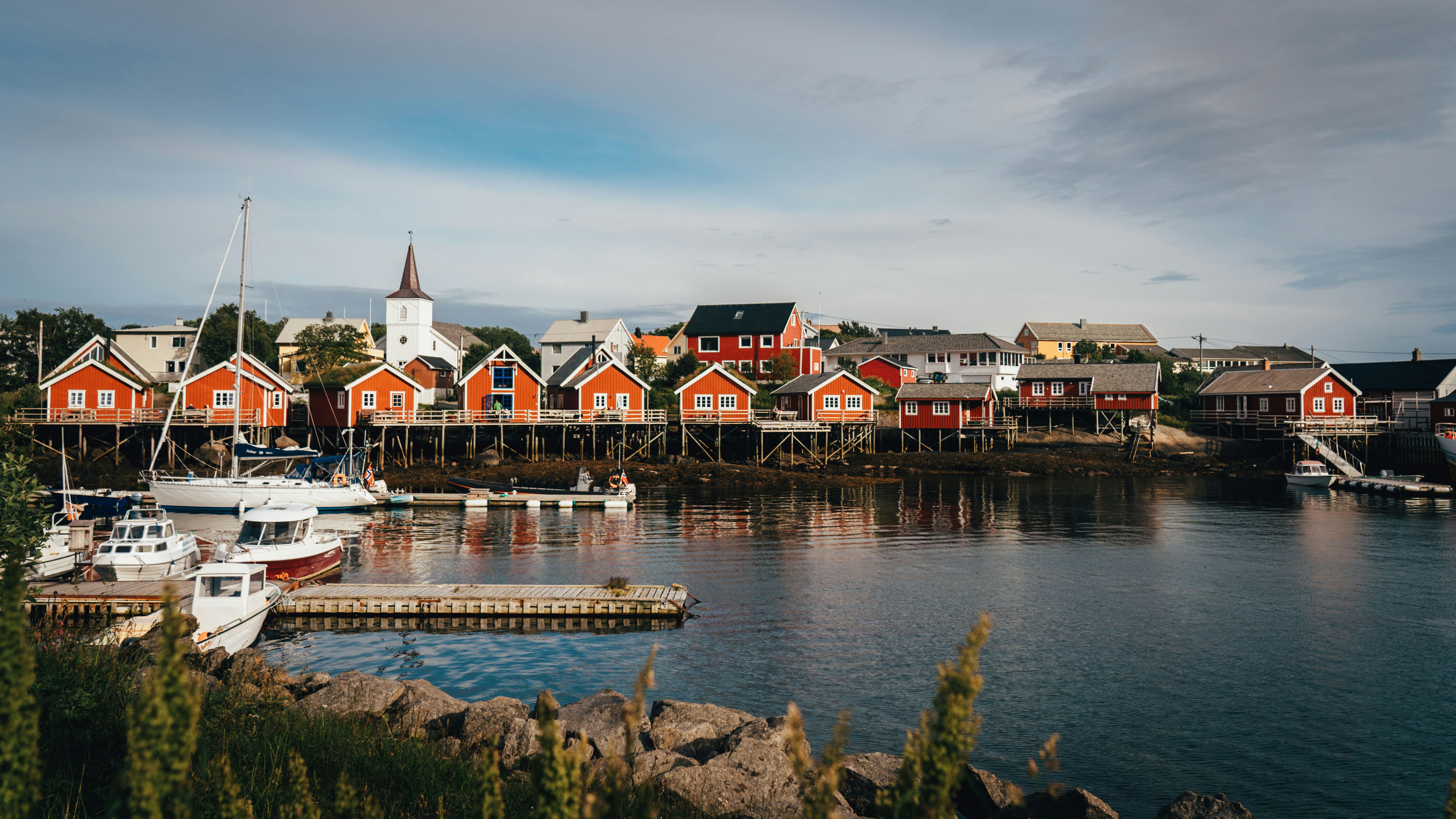 Red fishermen's cabins line the bay in Reine, Norway, with boats docked in calm waters beneath a cloudy sky.