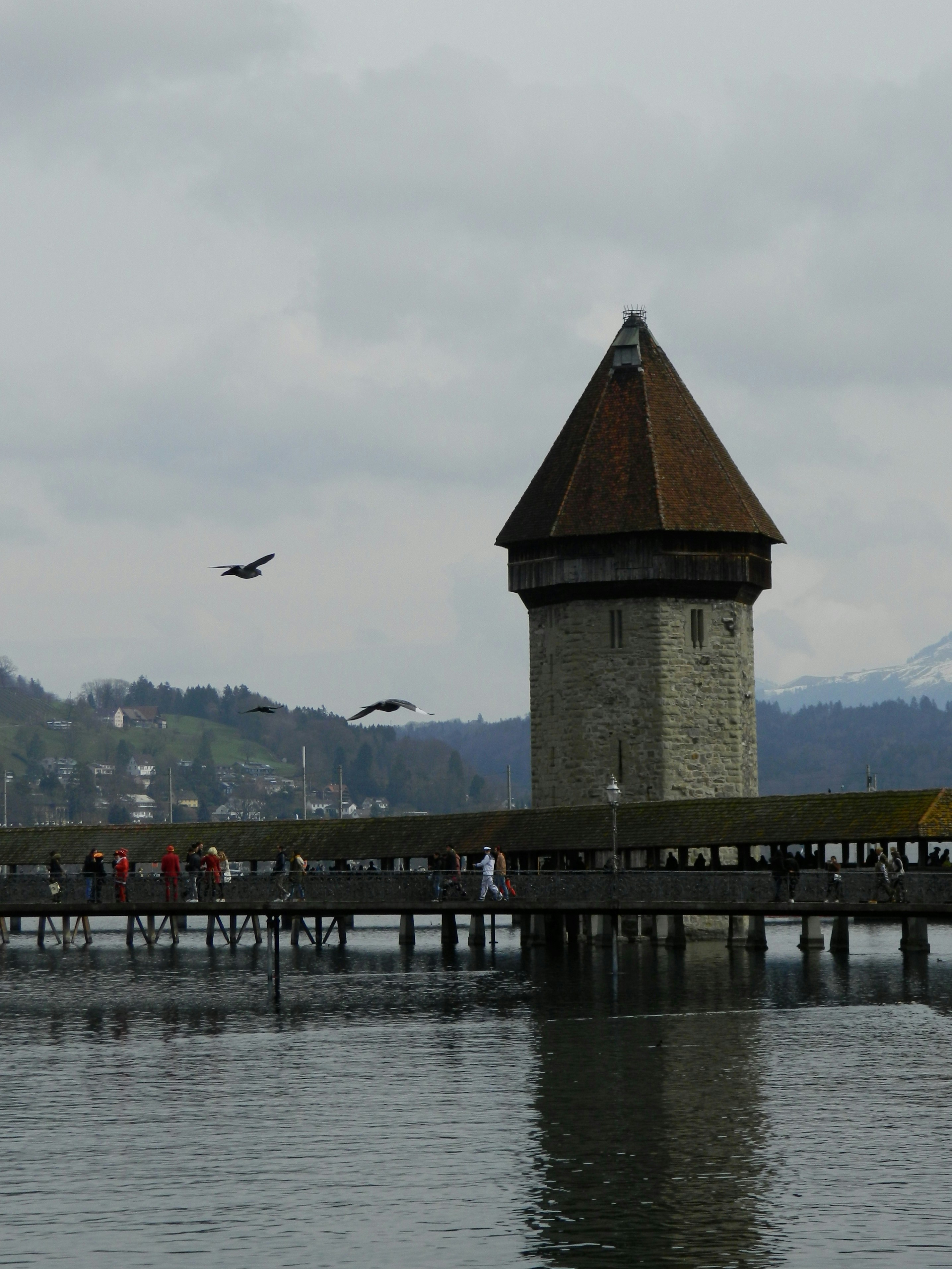 Historic tower beside a wooden pier with visitors strolling, under a cloudy sky. Birds fly overhead, adding life to the serene lakeside scene.