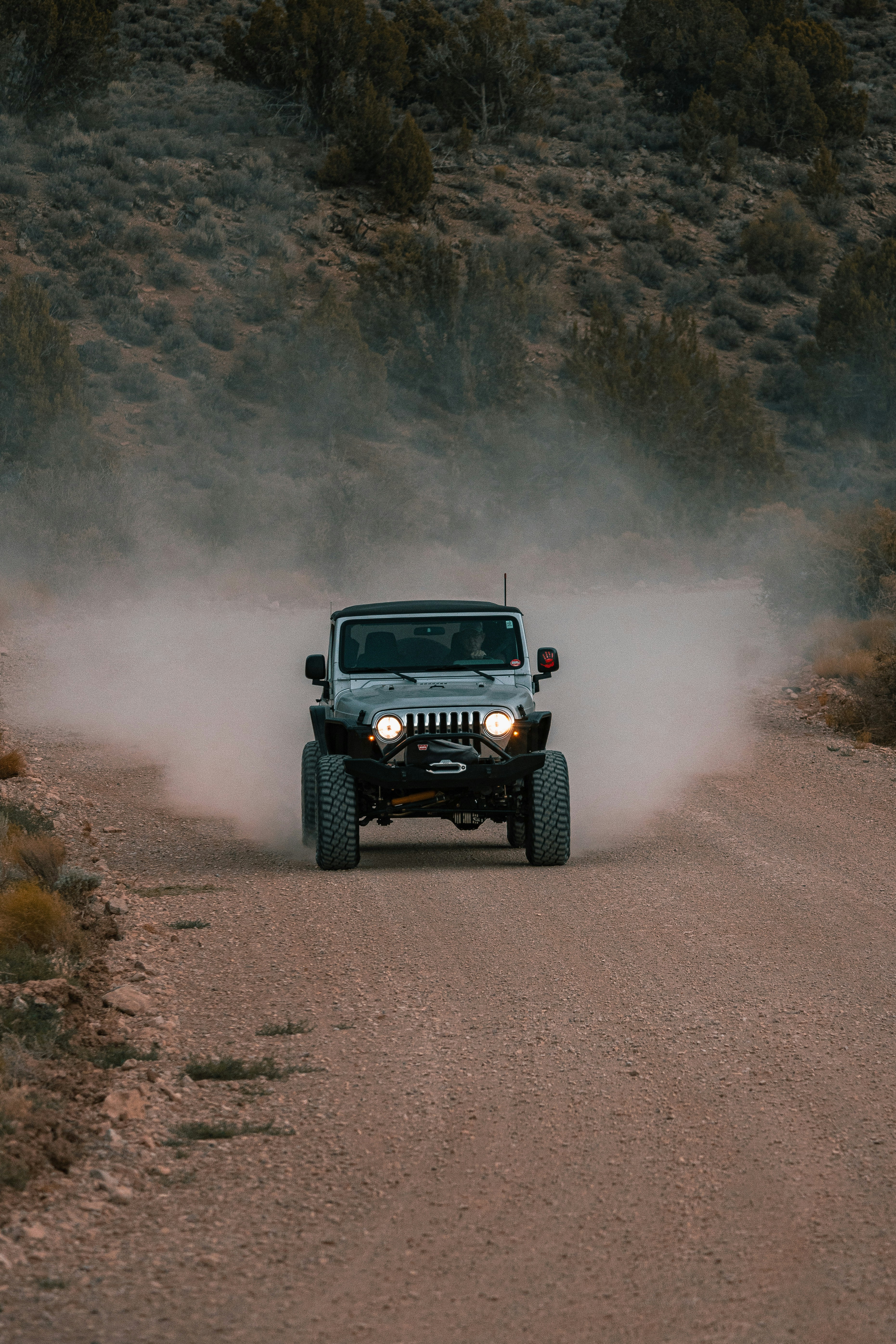 A rugged off-road vehicle kicking up dust on a gravel road surrounded by sparse vegetation and rocky terrain.