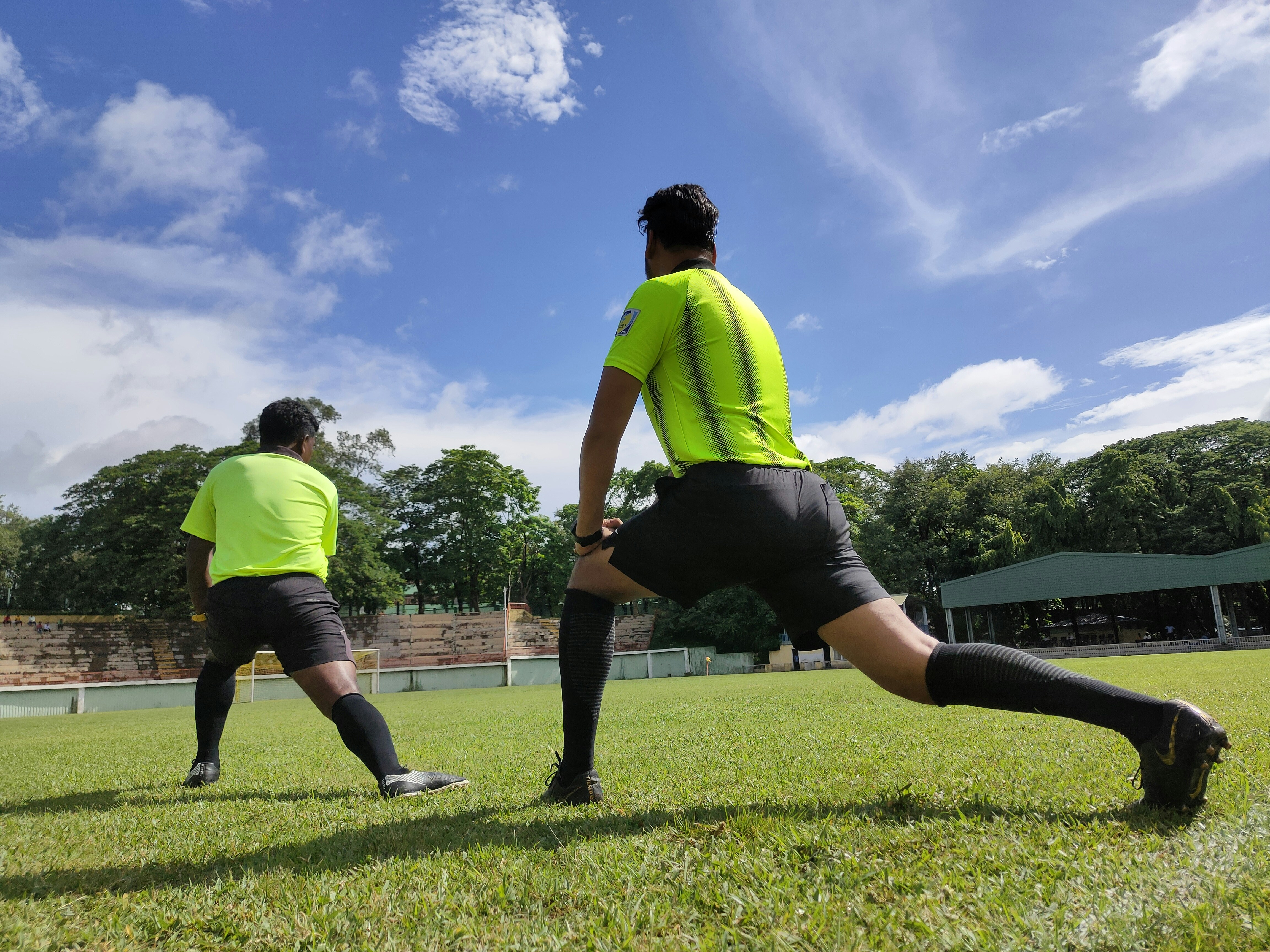 Two athletes in bright green shirts stretching on a lush field beneath a vibrant blue sky with scattered clouds.