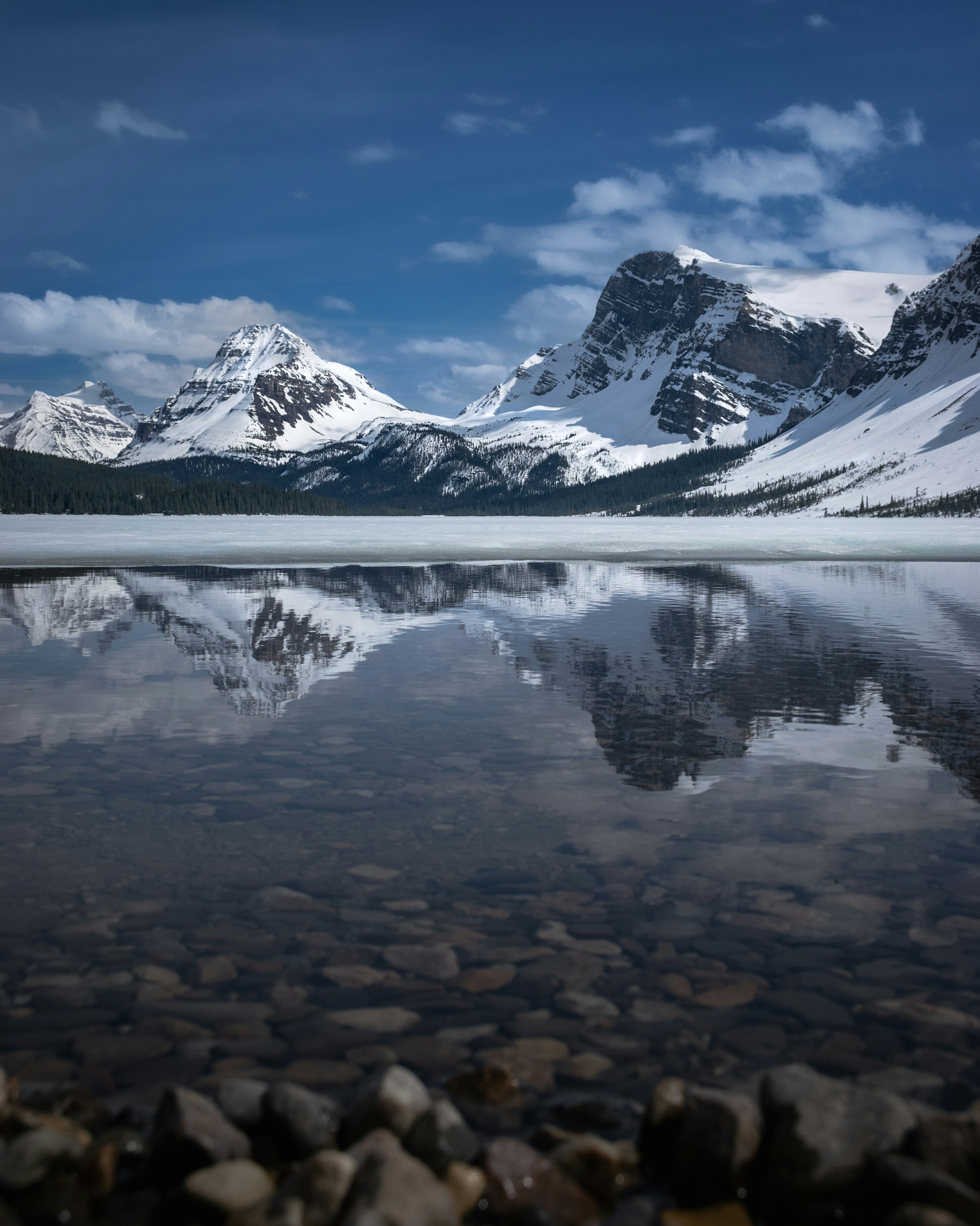 Low angle reflection at Bow Lake on a blue sky with clouds day. Banff, Canada, May/22. | a lake with snowy mountains in the background