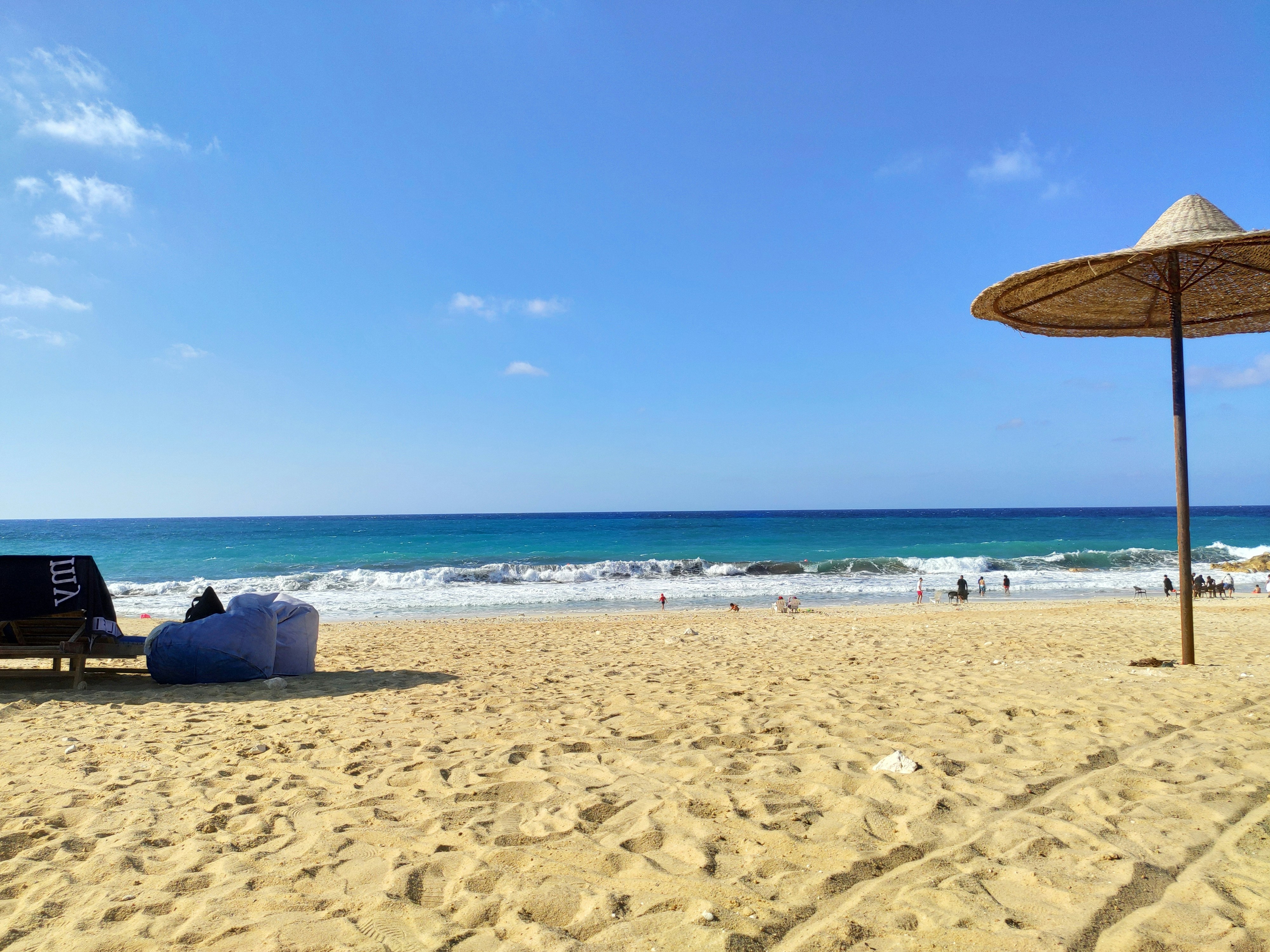 Golden sandy beach with a lone straw umbrella and distant figures in the blue sea.
