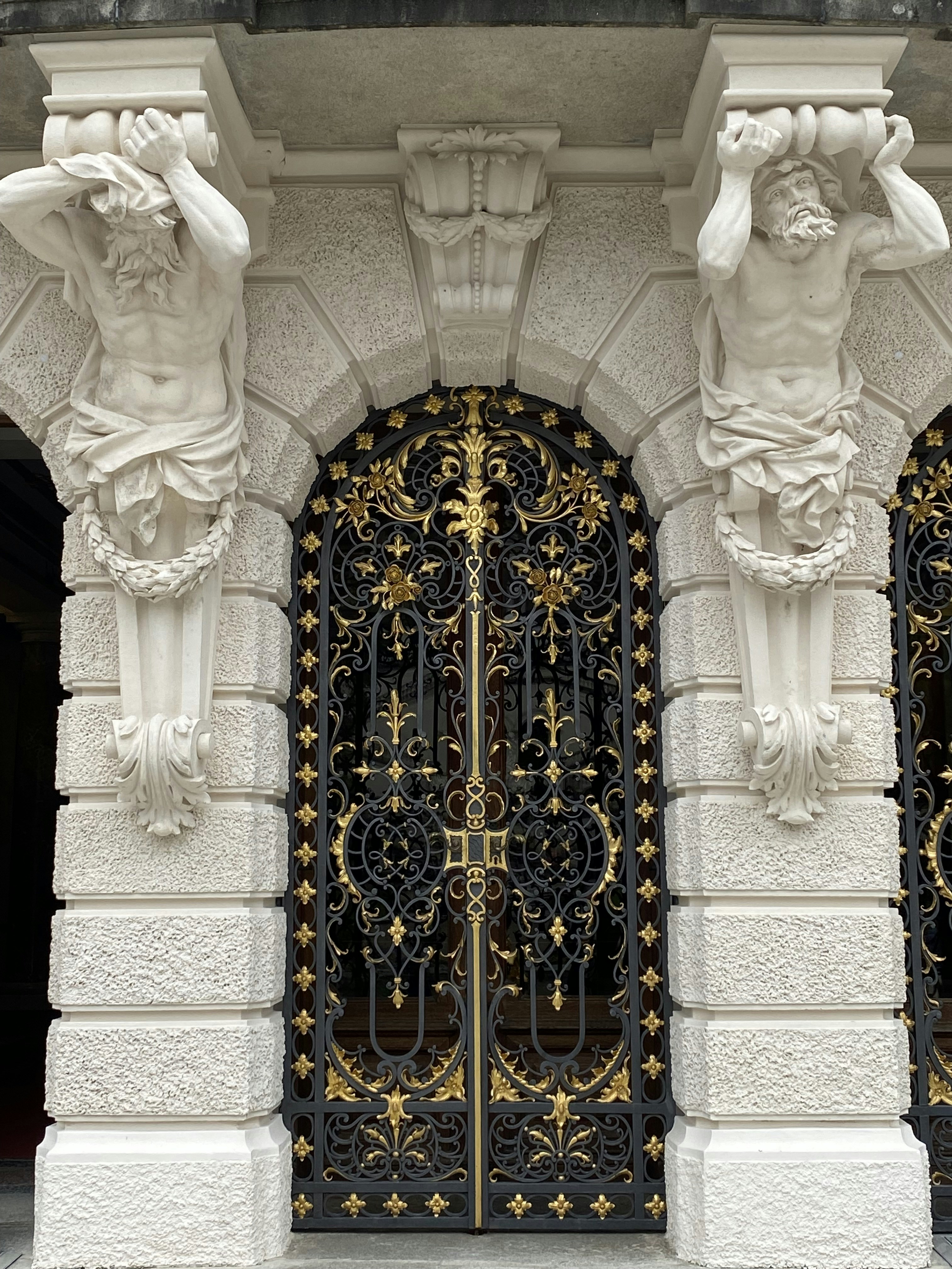 Intricate stone carvings of muscular figures flanking an ornate wrought iron door adorned with golden floral patterns.