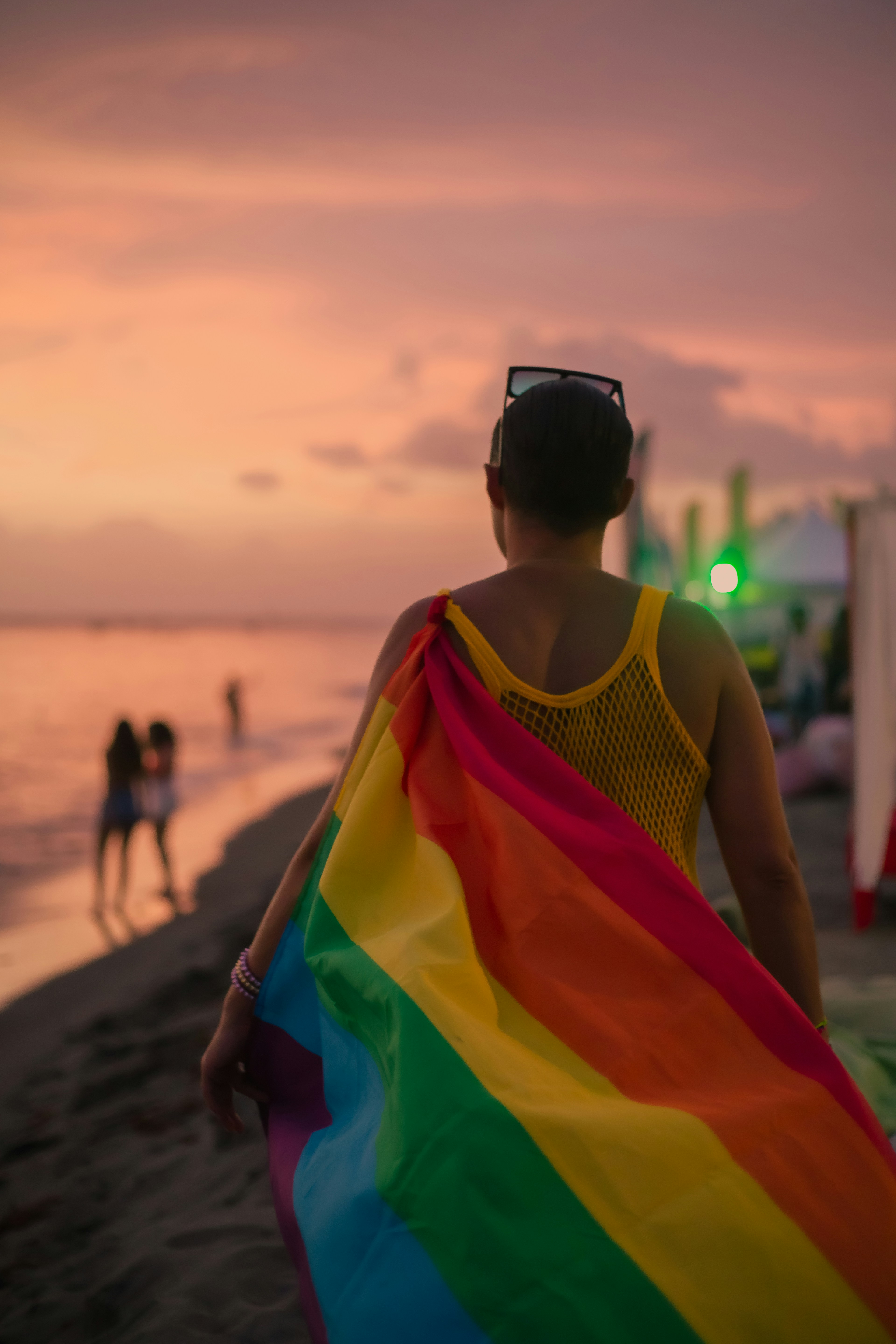 Individual draped in a rainbow flag walks along the beach at sunset, symbolizing pride and unity. Soft pastel hues fill the sky.