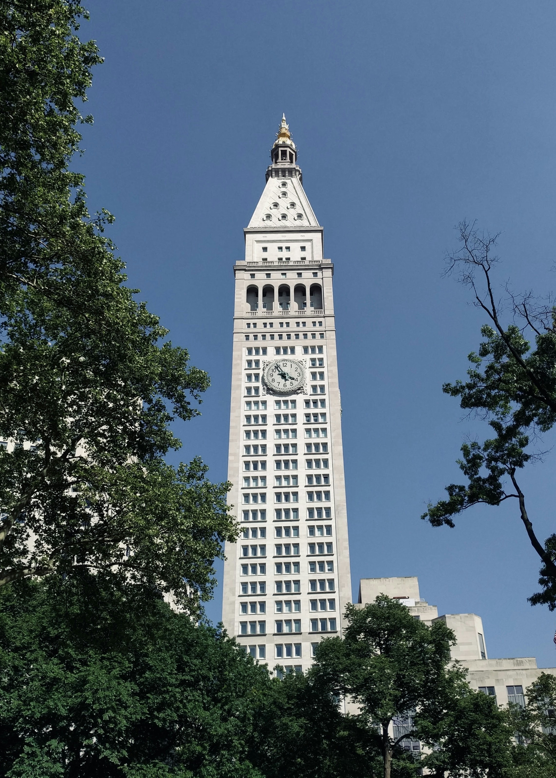 Tall clock-tower plaza ascent framed by leafy foreground and a vivid blue sky.