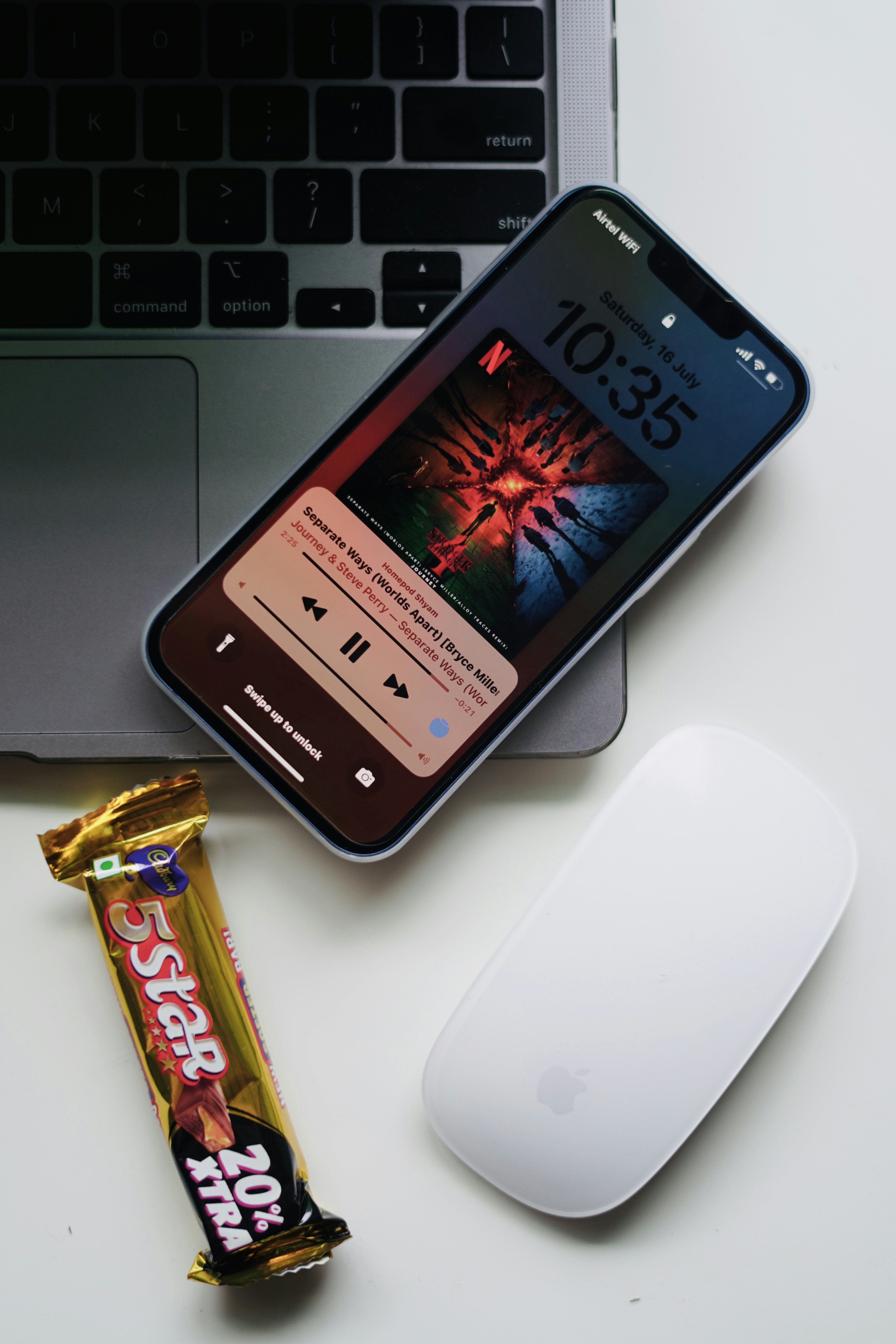 Smartphone resting on a laptop keyboard with a white wireless mouse nearby and a chocolate bar in the foreground. The scene highlights a modern workspace setup and a music/comms moment.