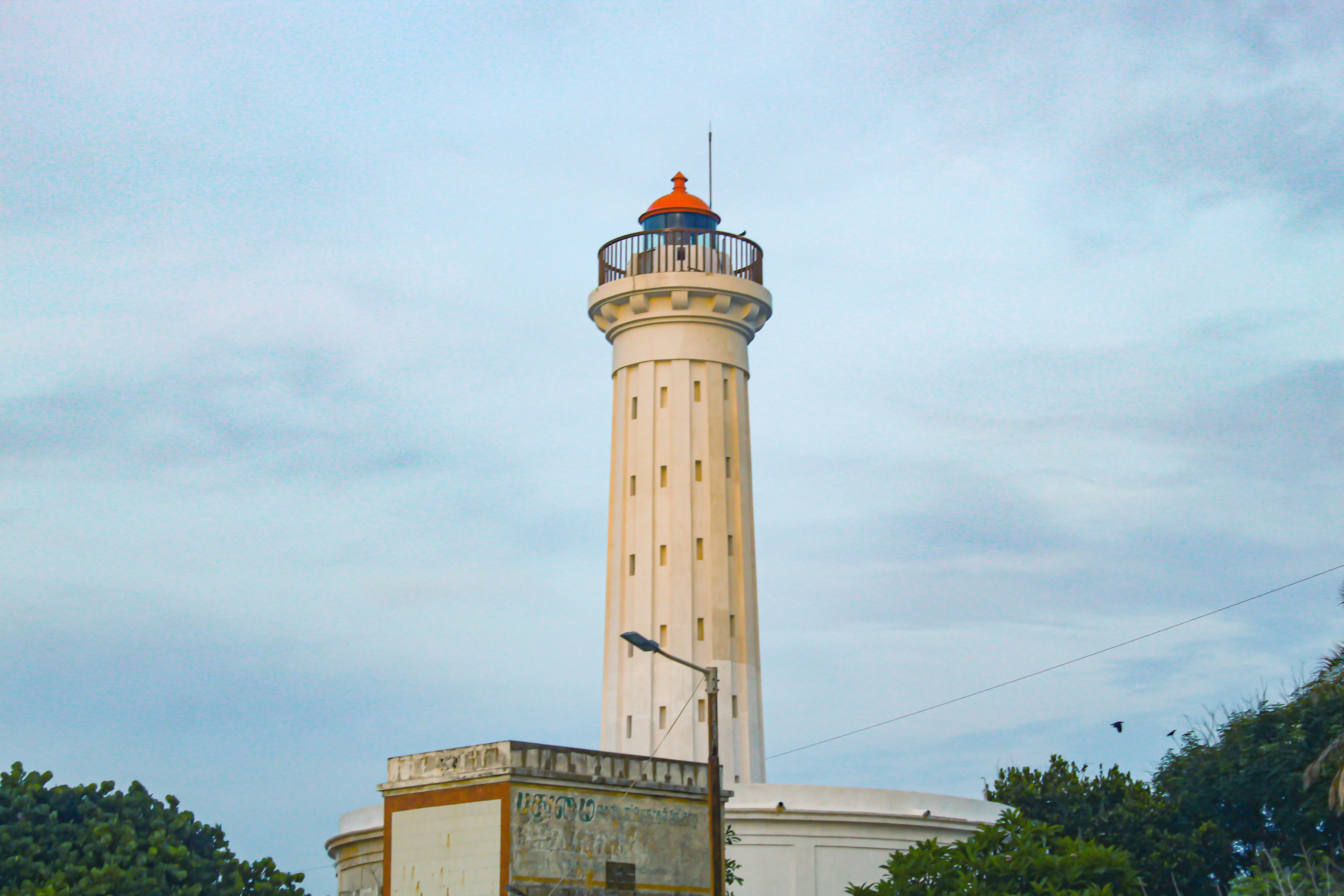 A historic lighthouse standing tall against a cloudy sky, surrounded by lush greenery and urban structures.