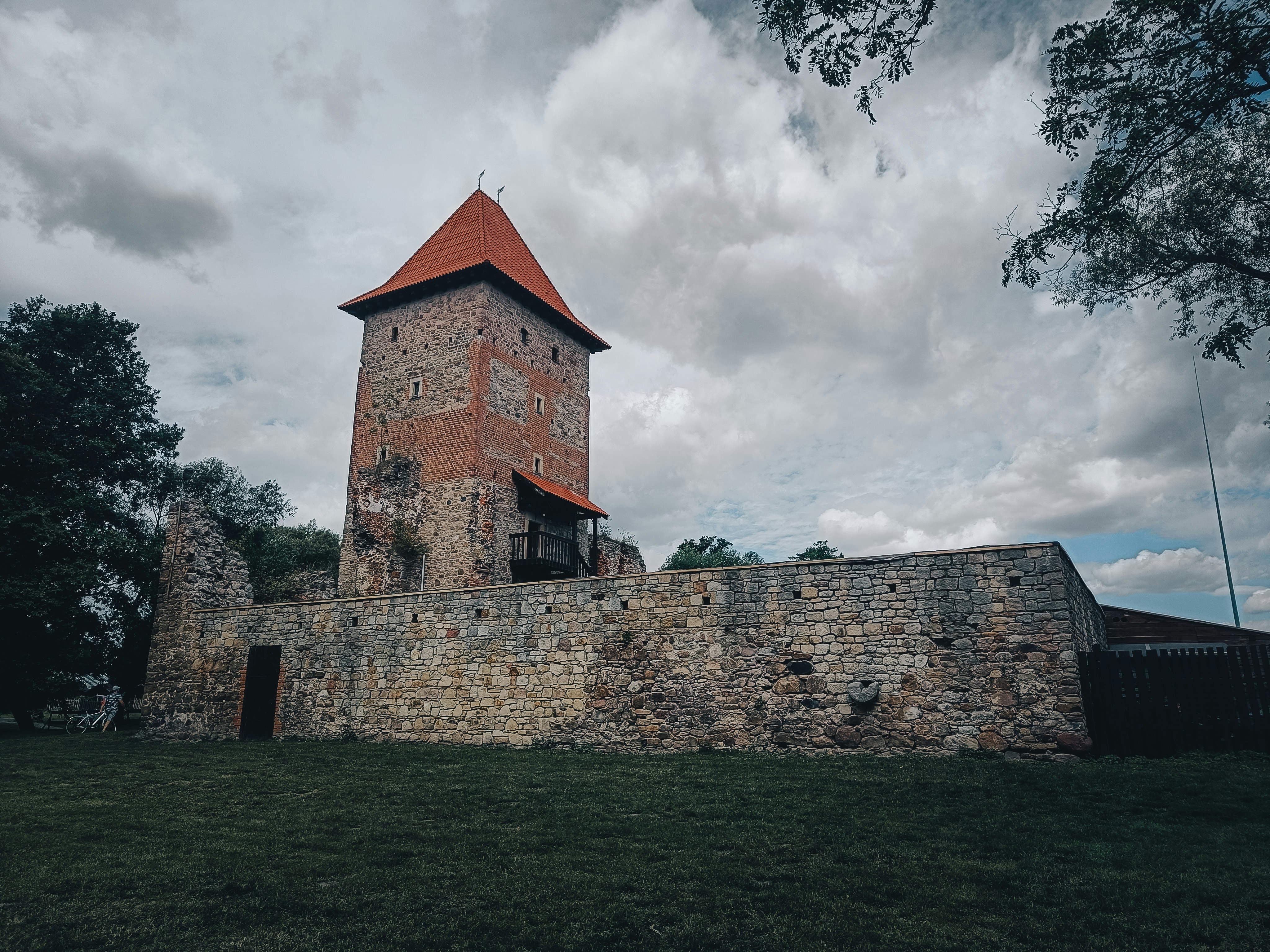 Ancient stone tower with a distinctive red roof stands amidst lush greenery and cloudy skies, showcasing remnants of a bygone era.