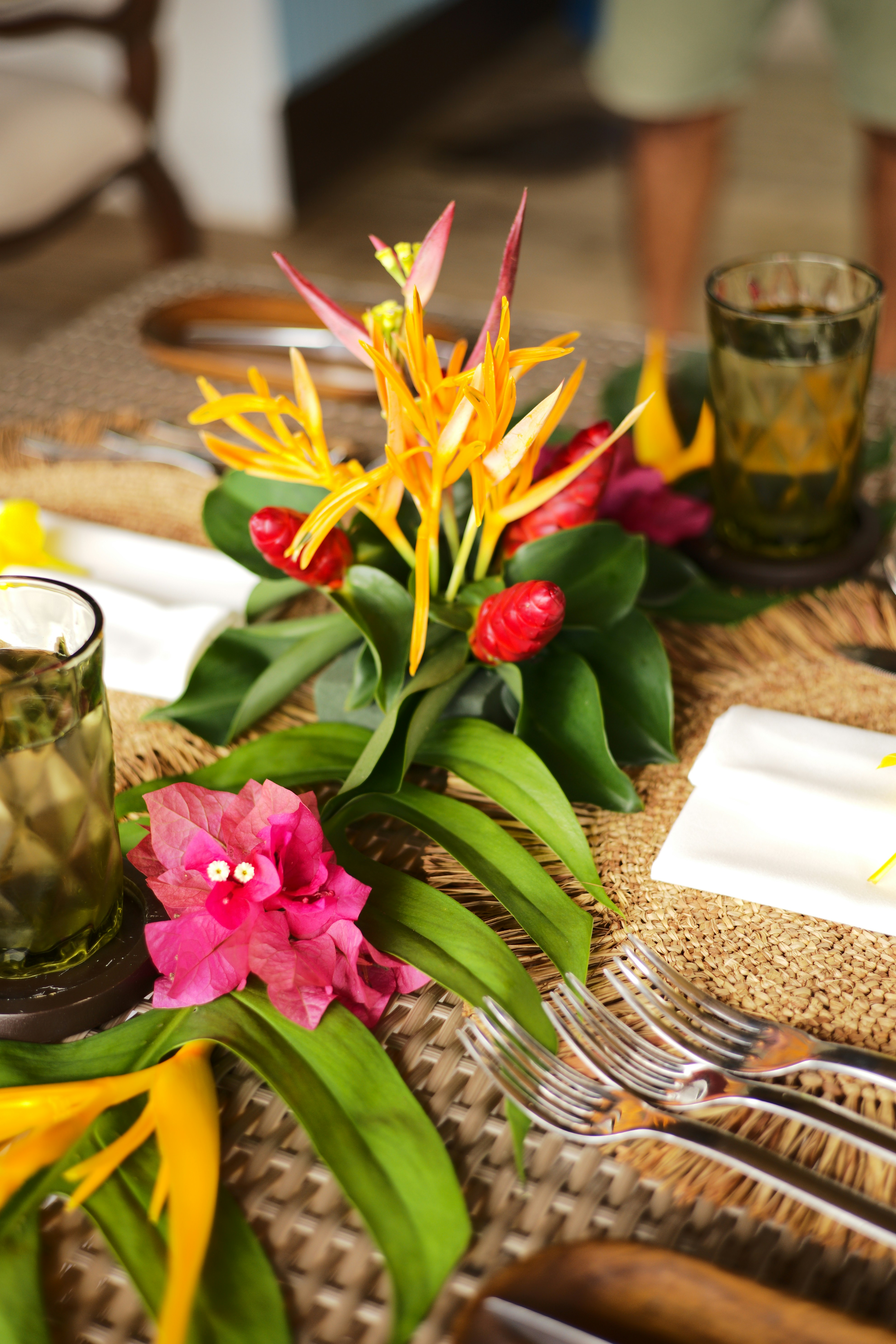 Tropical flower centerpiece on a woven dining table with glass tumblers and folded napkins.