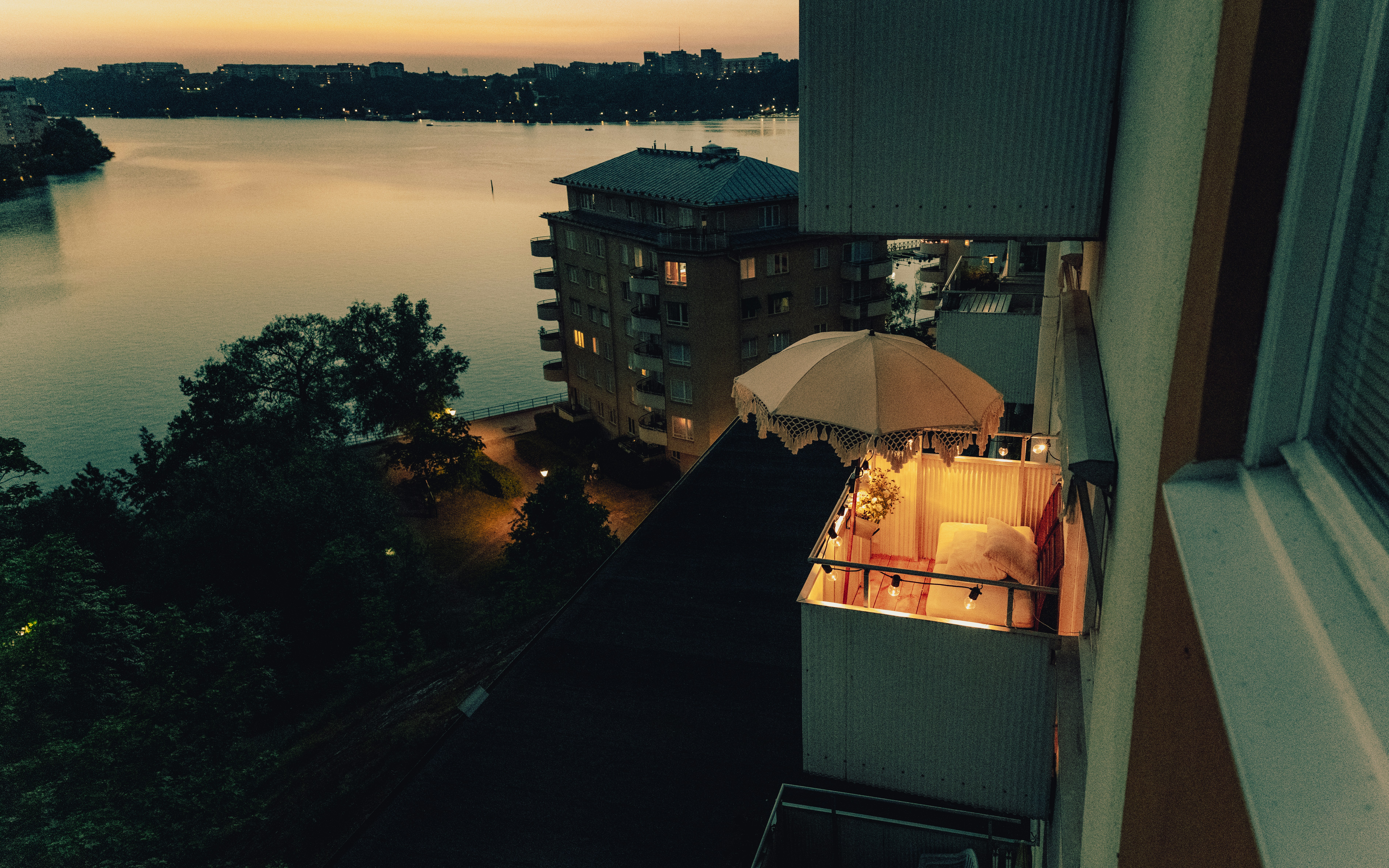 Cozy balcony adorned with a parasol overlooking a tranquil lake at dusk, illuminated by warm lights. The scene captures a peaceful urban retreat.
