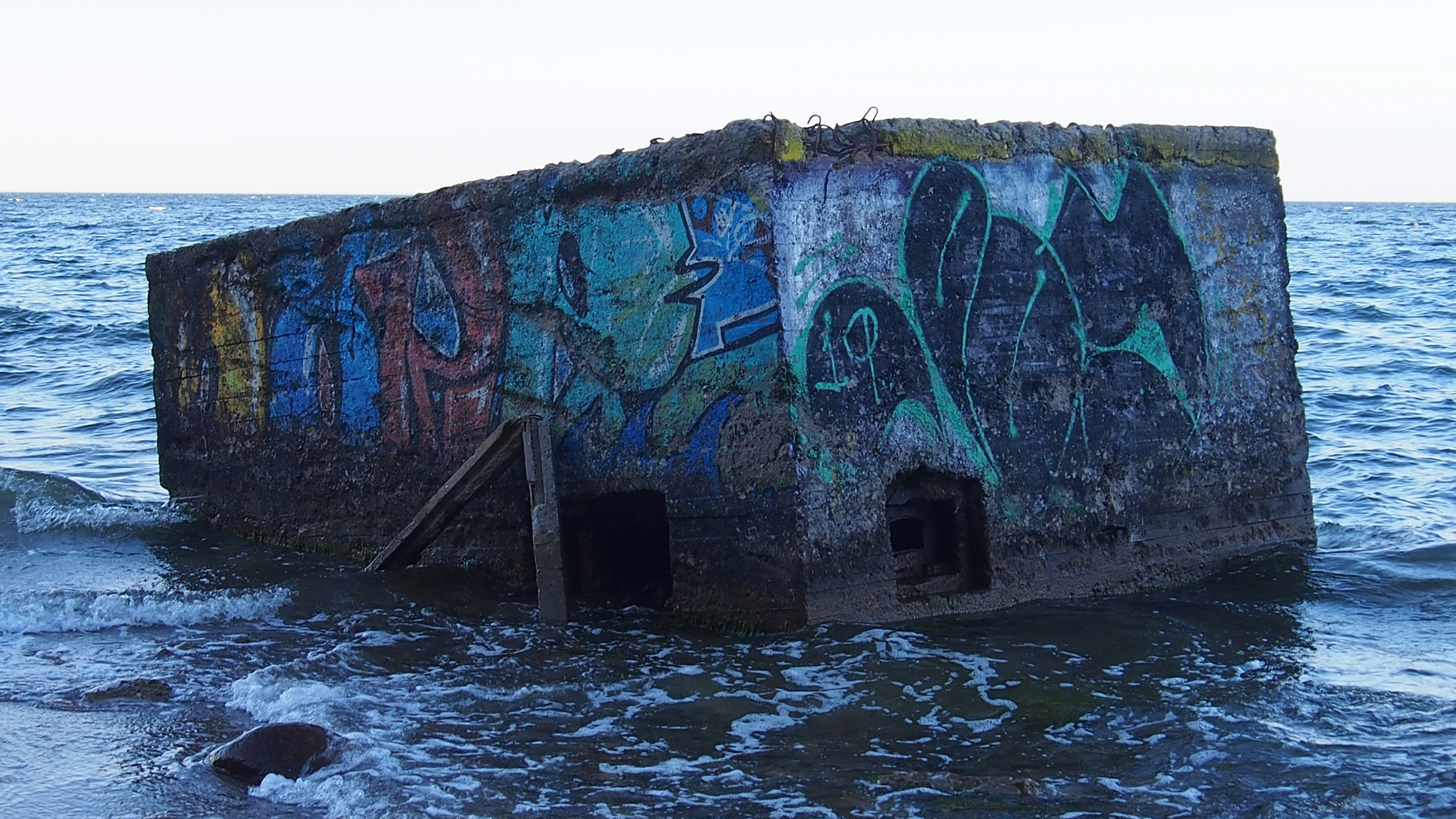 Weathered concrete structure adorned with vibrant graffiti, partially submerged in the ocean waves. The scene captures a blend of urban art and nature's reclaiming force.