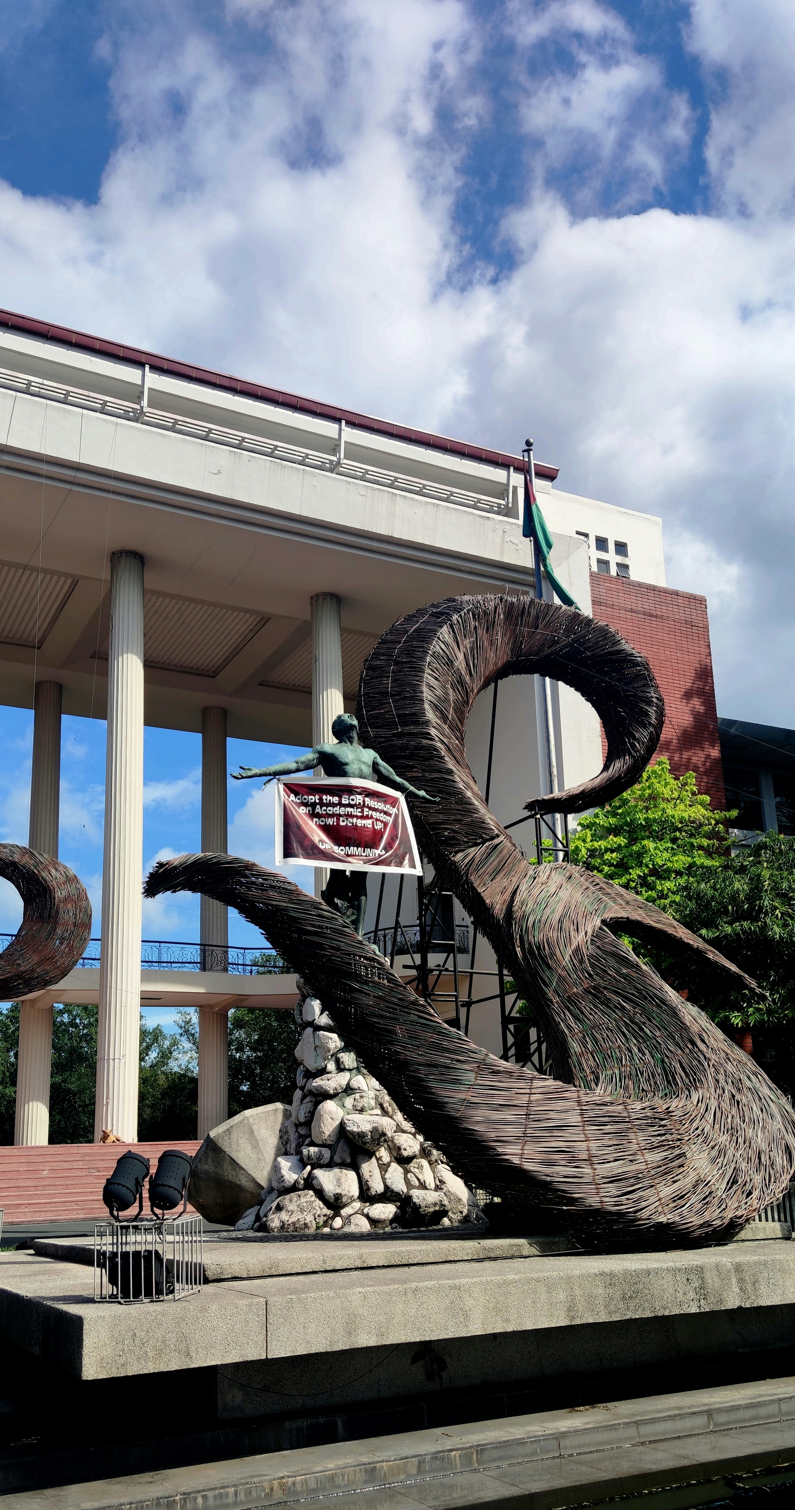Intricate sculpture of intertwined vines and stone, set against a backdrop of classical architecture and vibrant greenery.