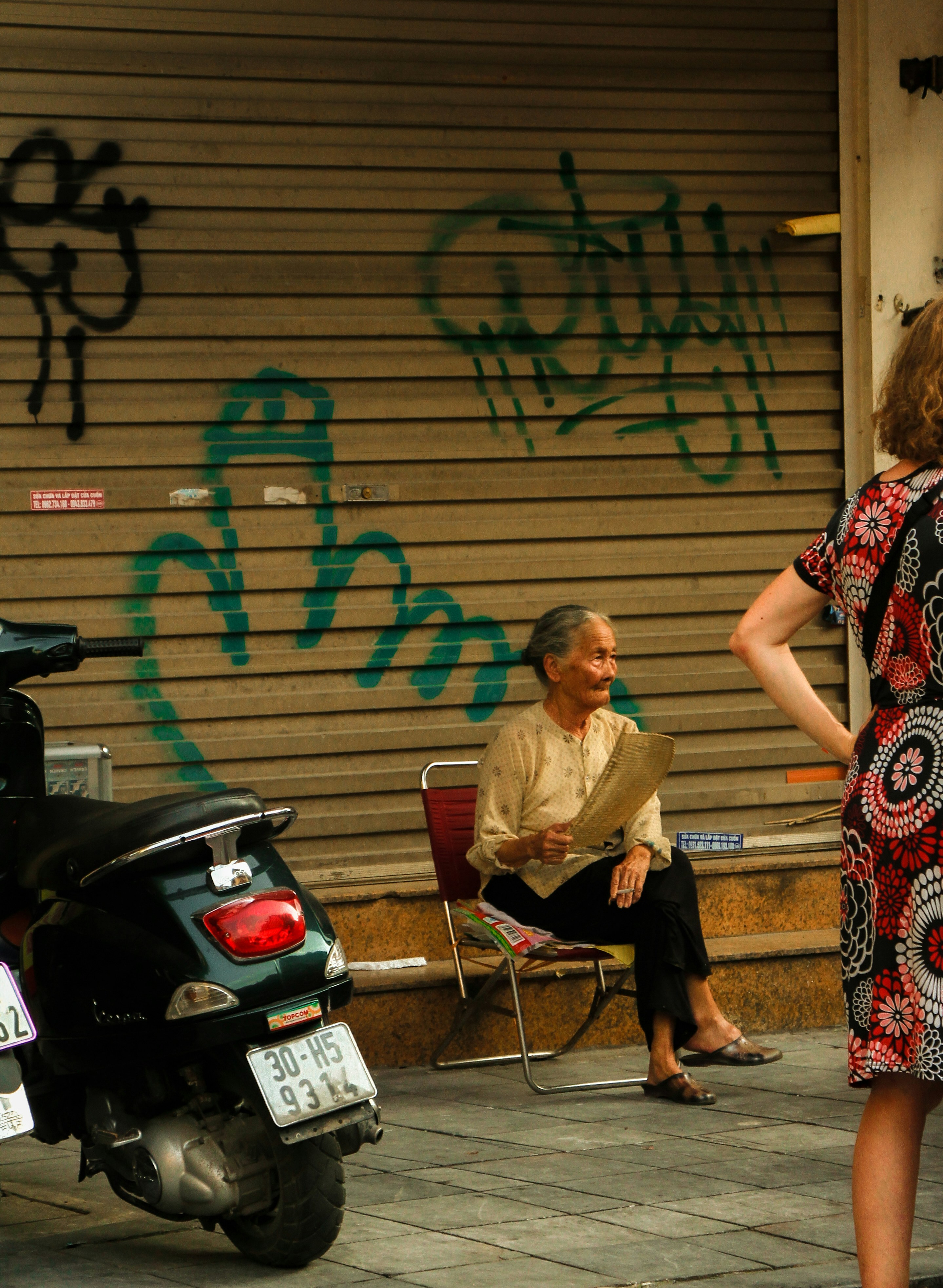 Elderly woman seated on a chair, fanning herself, against a backdrop of urban graffiti. A motorcycle is parked nearby.