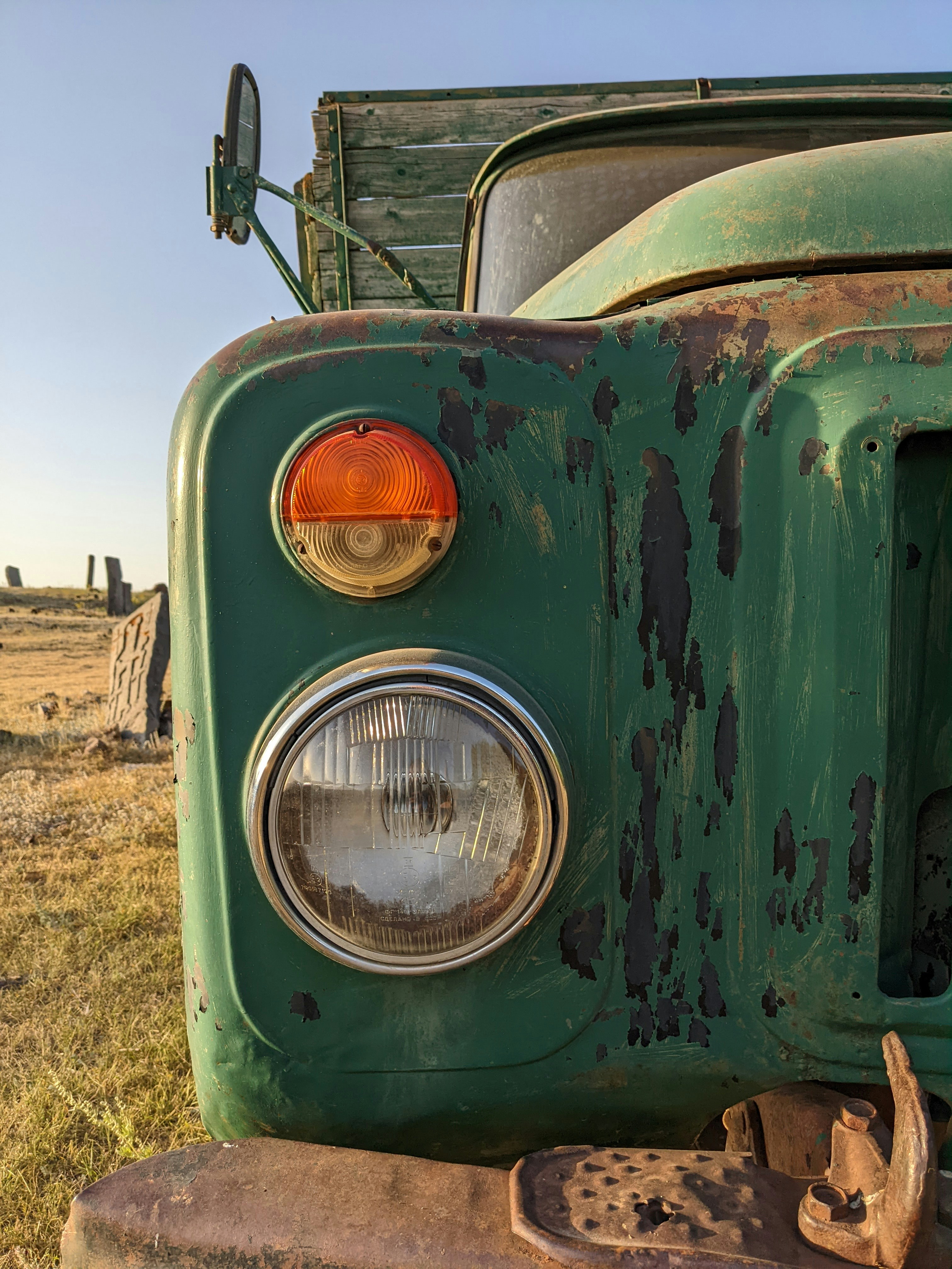 Close-up of a rusted green vintage truck front with a round headlight and orange signal lamp, set in a sunlit field.