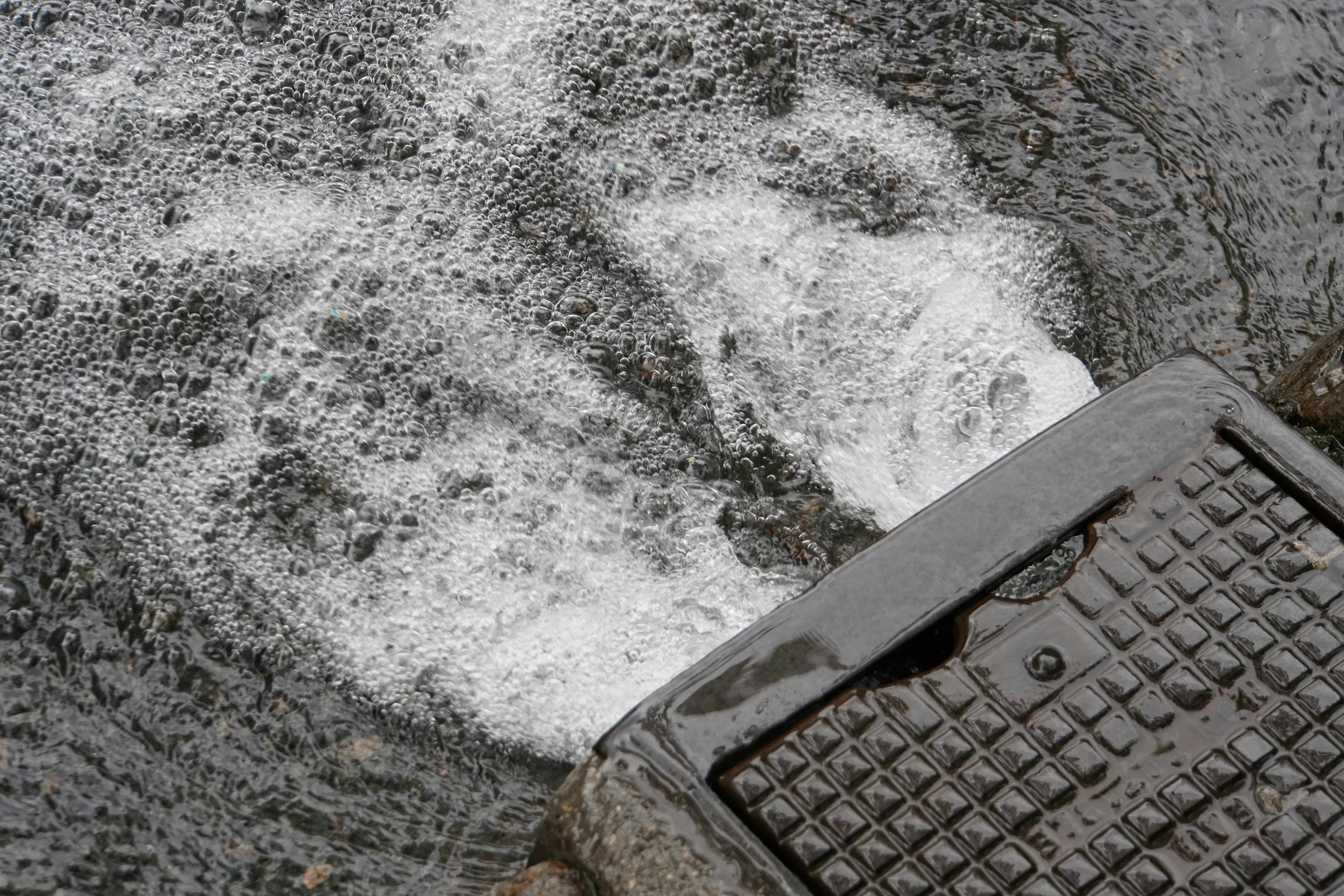 Foamy water flowing over a textured grate in an urban setting, highlighting the interplay between nature and infrastructure.
