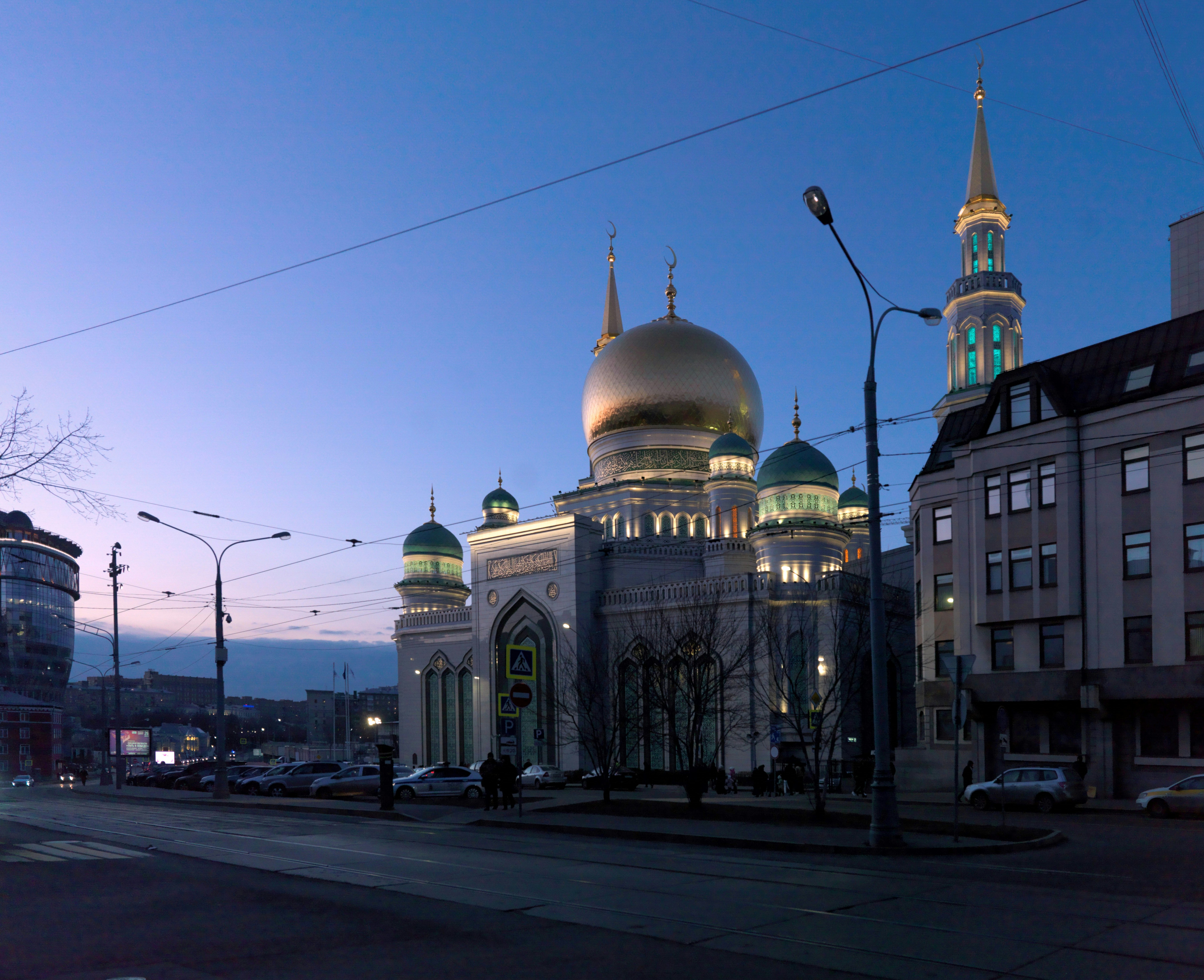 Dusk-lit street scene featuring a grand mosque with a gold central dome and green cupolas, illuminated by streetlights and framed by surrounding urban buildings.