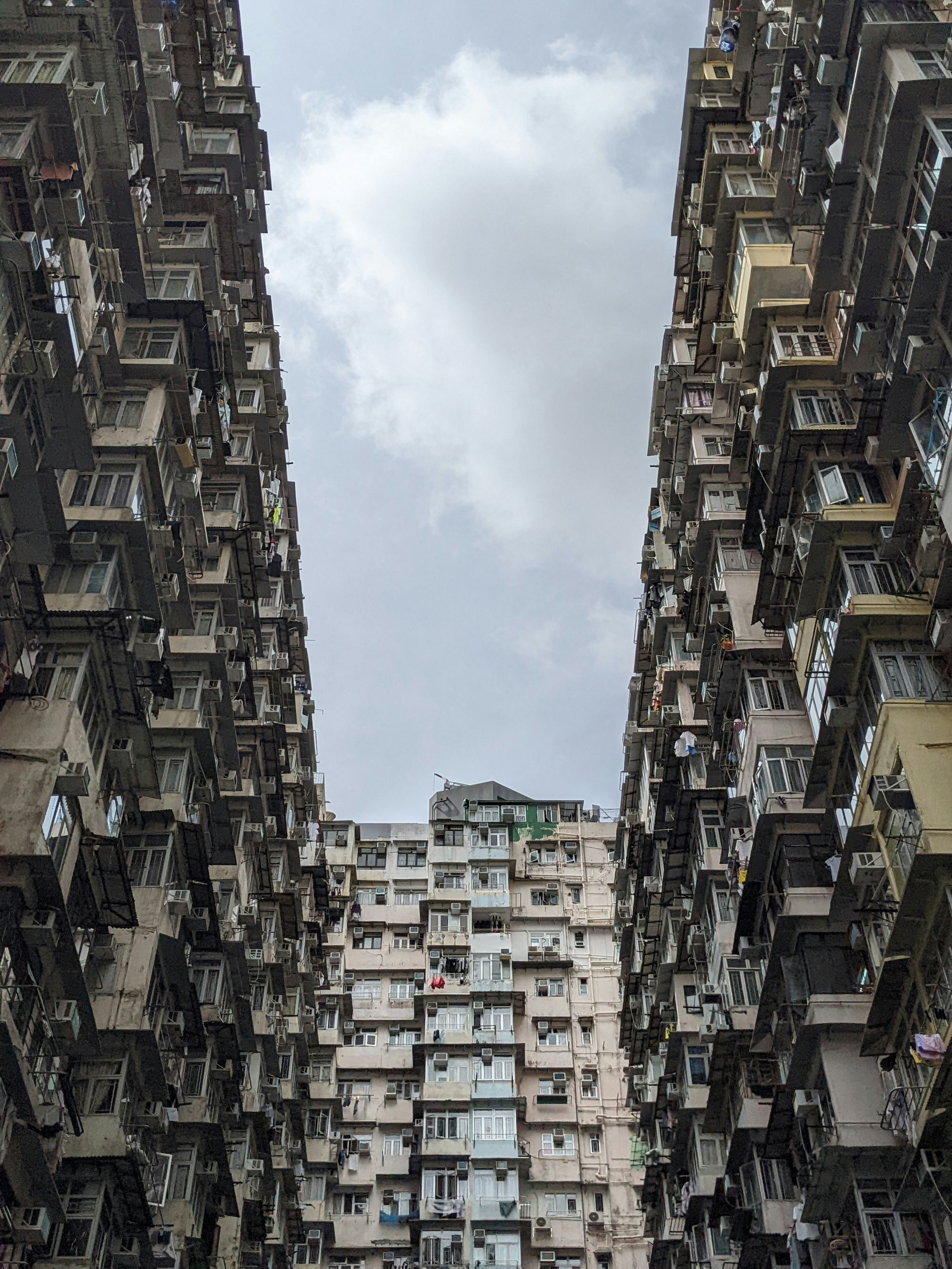 Vertical view of densely packed apartment buildings creating a narrow corridor, with clouds visible above. The architecture reflects urban living dynamics.