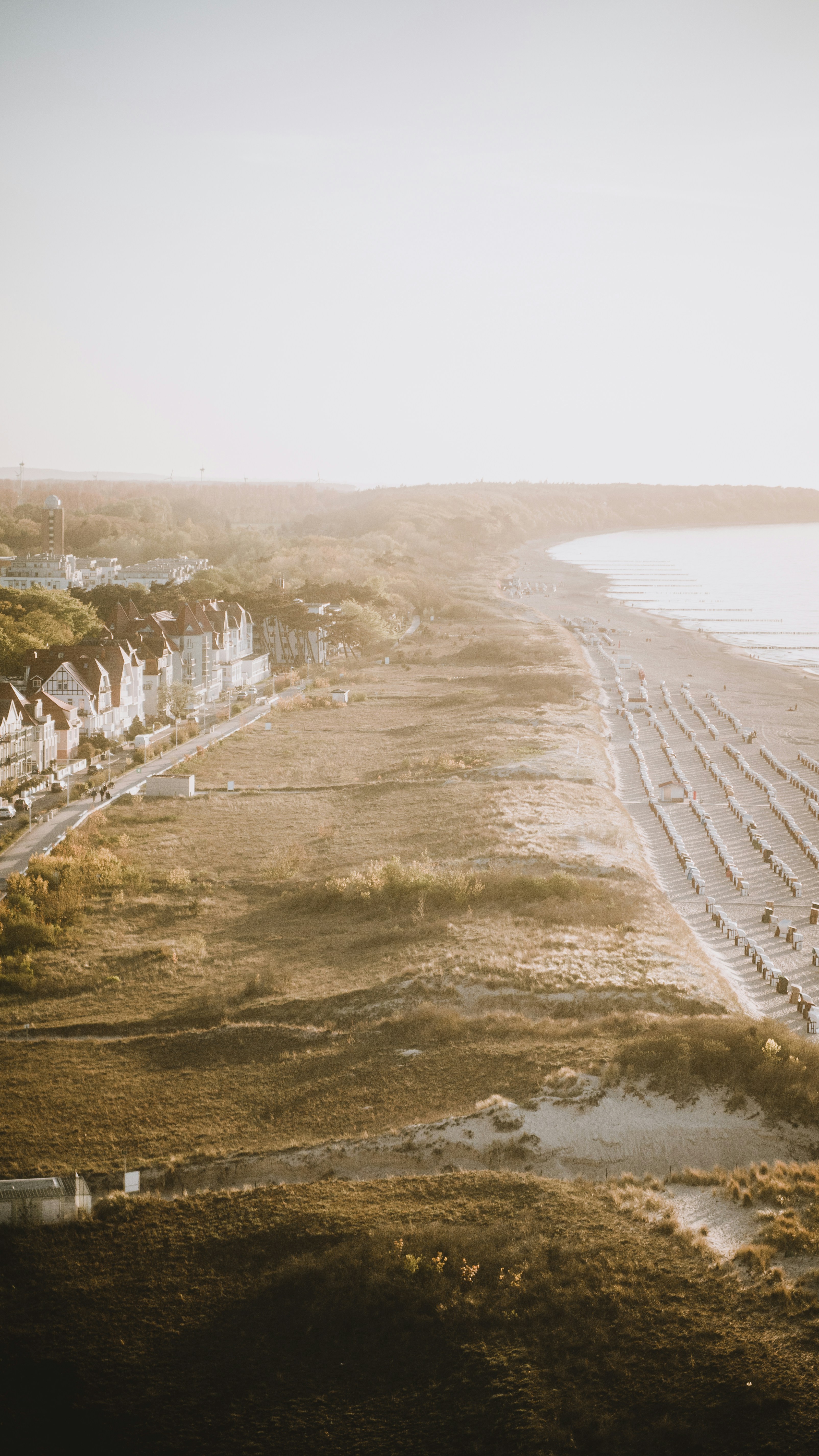 A peaceful beach scene with gentle waves lapping at the shore, framed by distant coastal homes and lush greenery. The soft light creates a serene atmosphere.