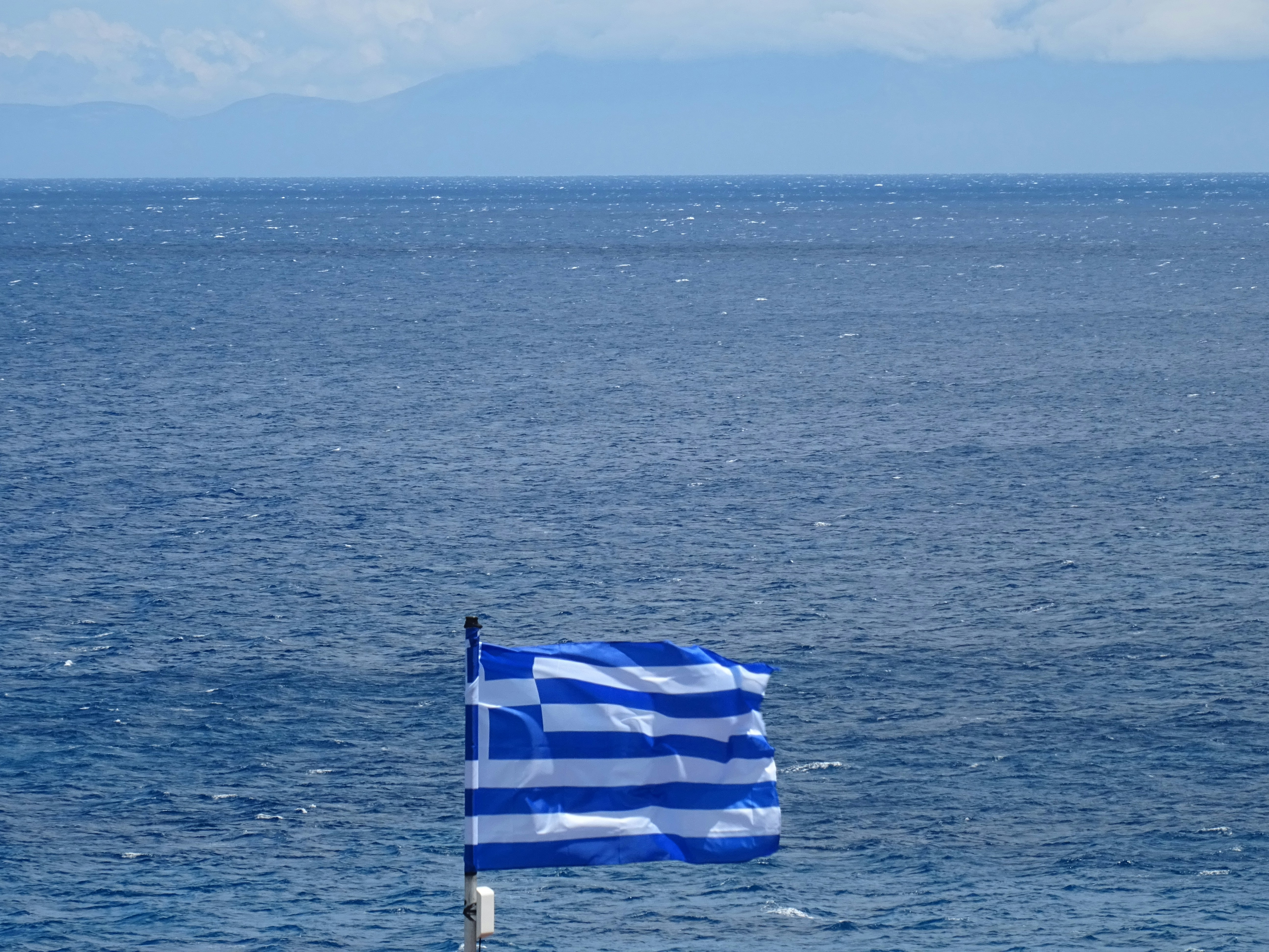 A Greek flag on a flagpole rises above a calm blue sea. The expansive horizon emphasizes the lone emblem against the ocean.