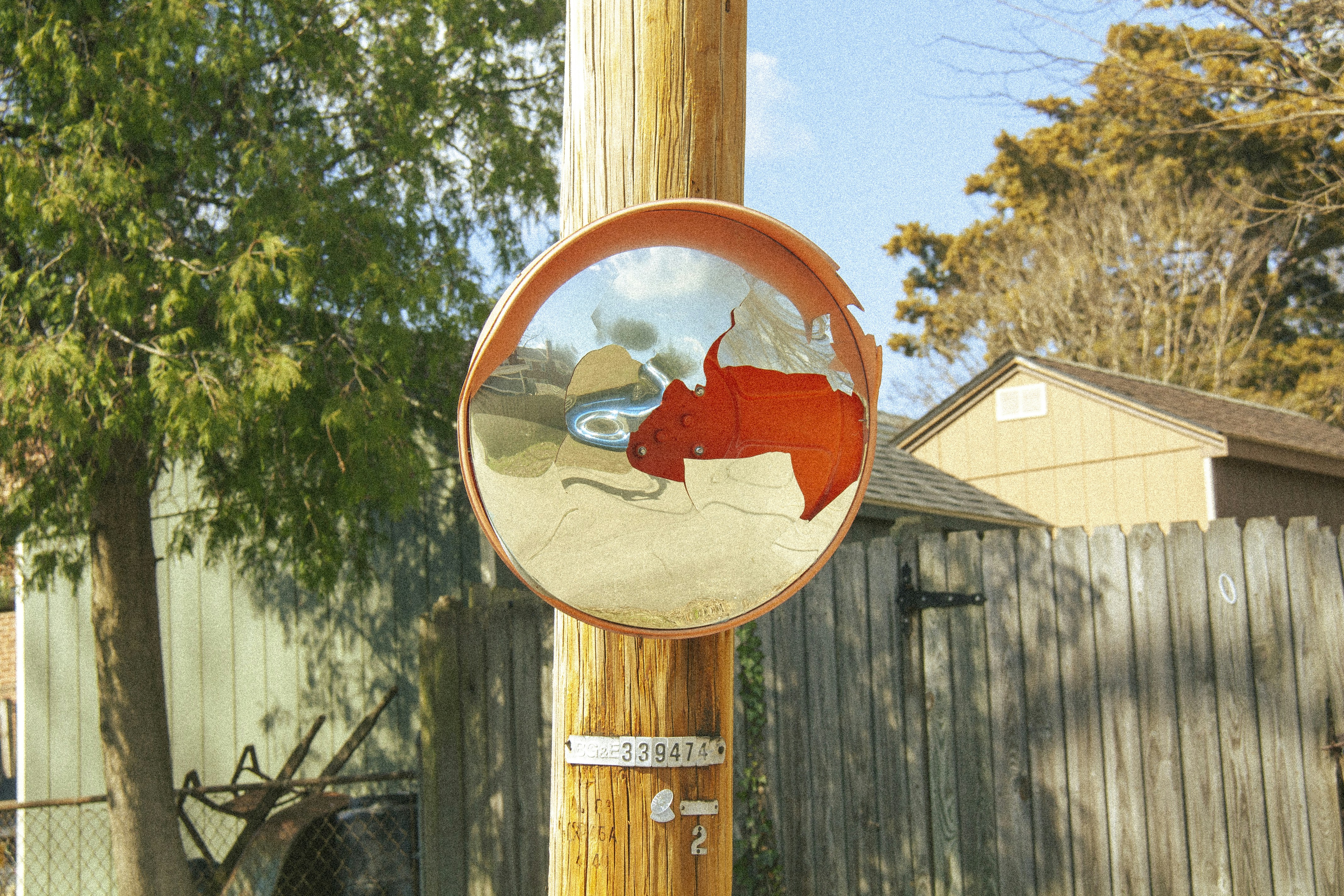 A convex mirror mounted on a wooden post reflects a vibrant red object and a glimpse of the surroundings, capturing a moment of urban life.