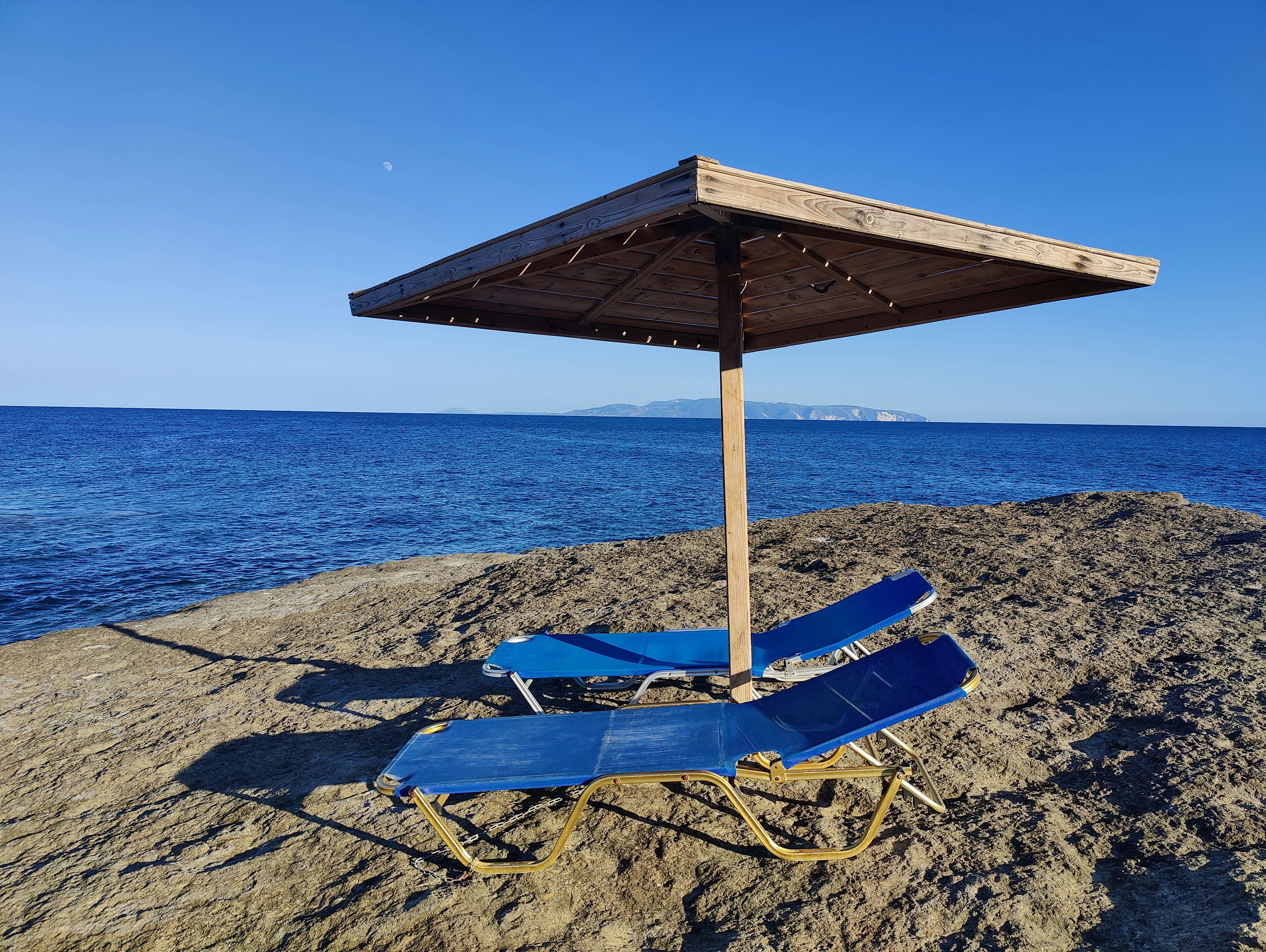 A wooden canopy shades two blue lounge chairs on a rocky shore, overlooking a tranquil sea. The distant island adds depth to the coastal scene.