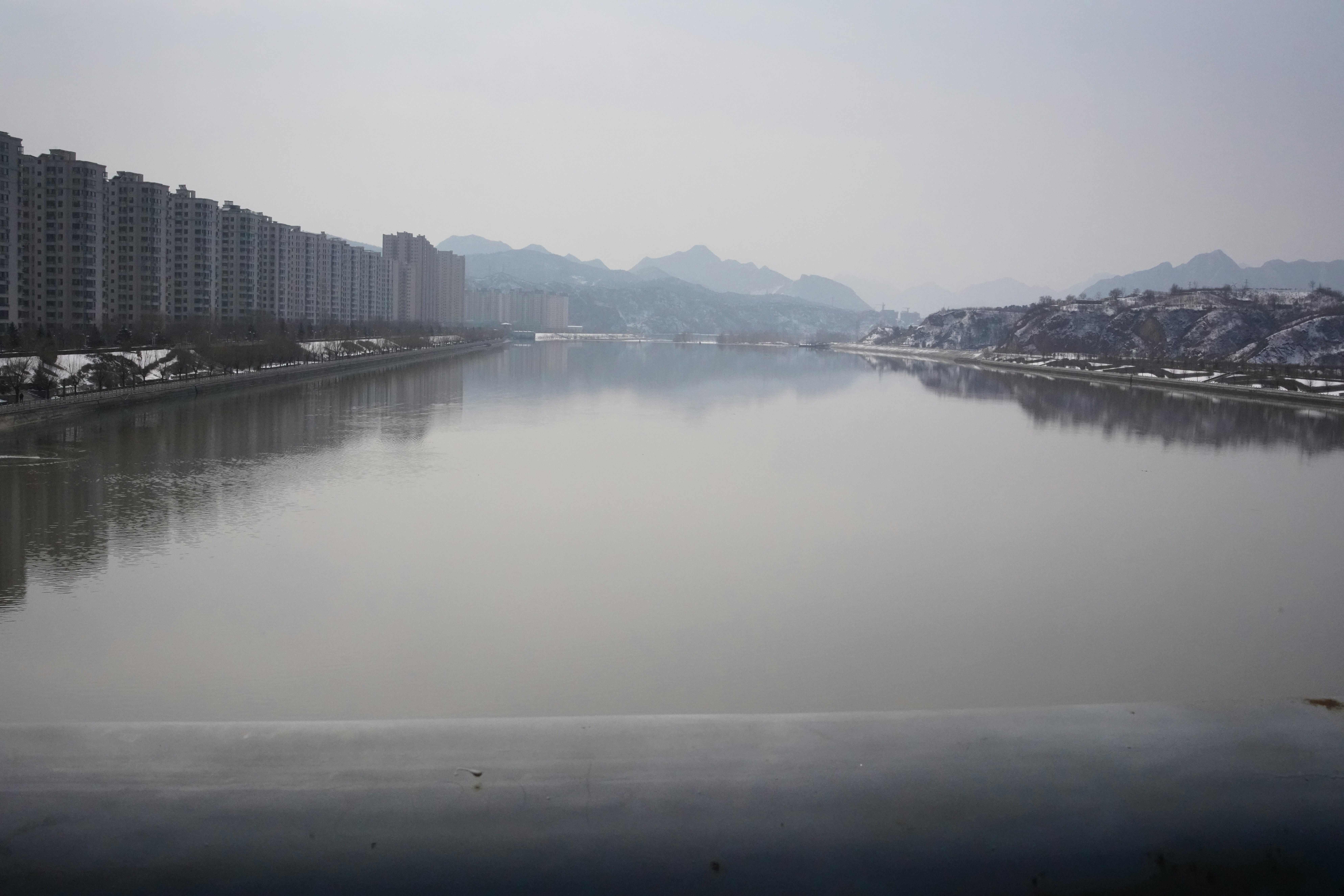 Calm river reflecting cityscape and mountains under a hazy sky. The stillness captures the essence of tranquility in an urban environment.