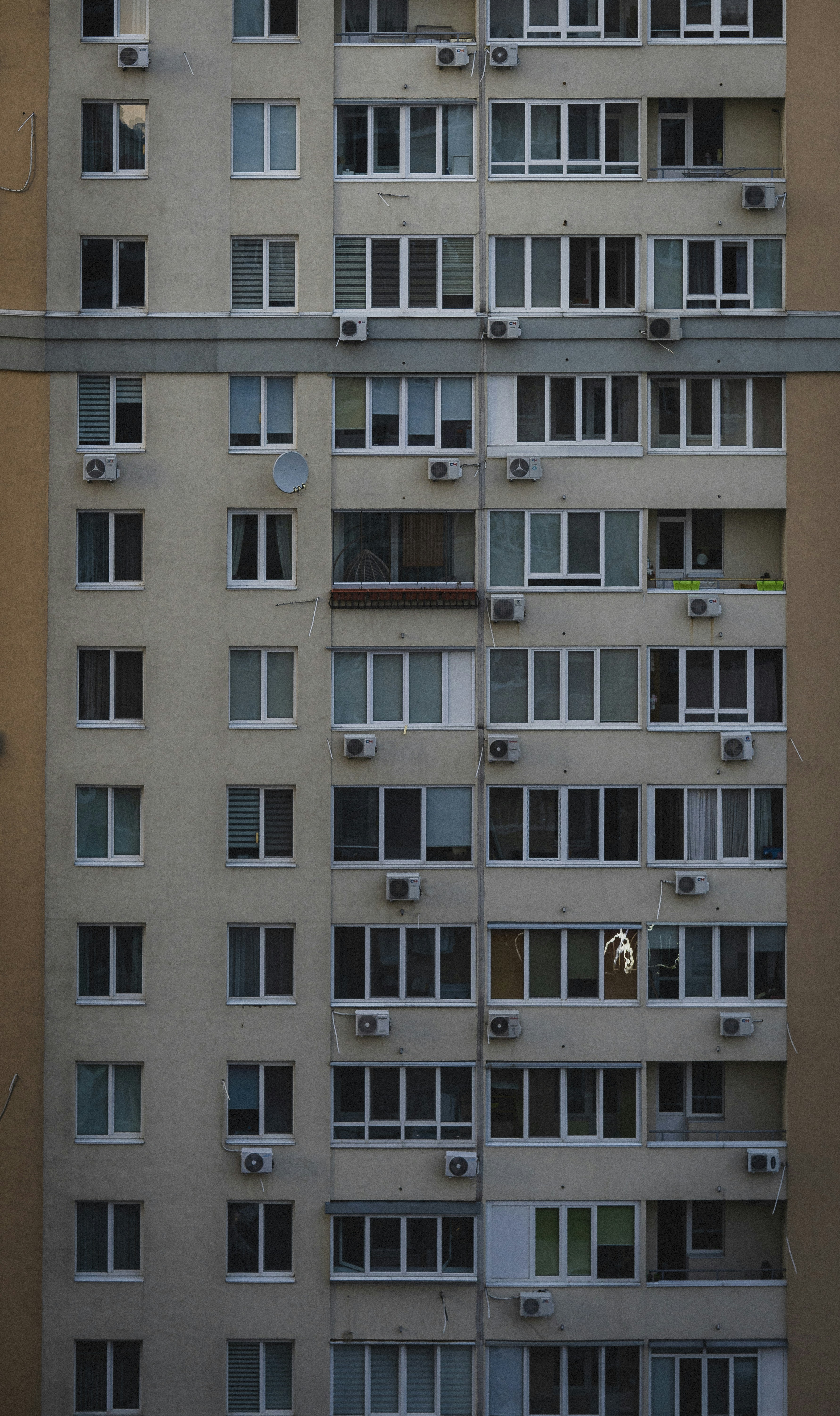 A high-rise apartment facade showcasing a grid of windows, air conditioning units, and a lone figure in the light. The scene reflects urban life and architectural patterns.