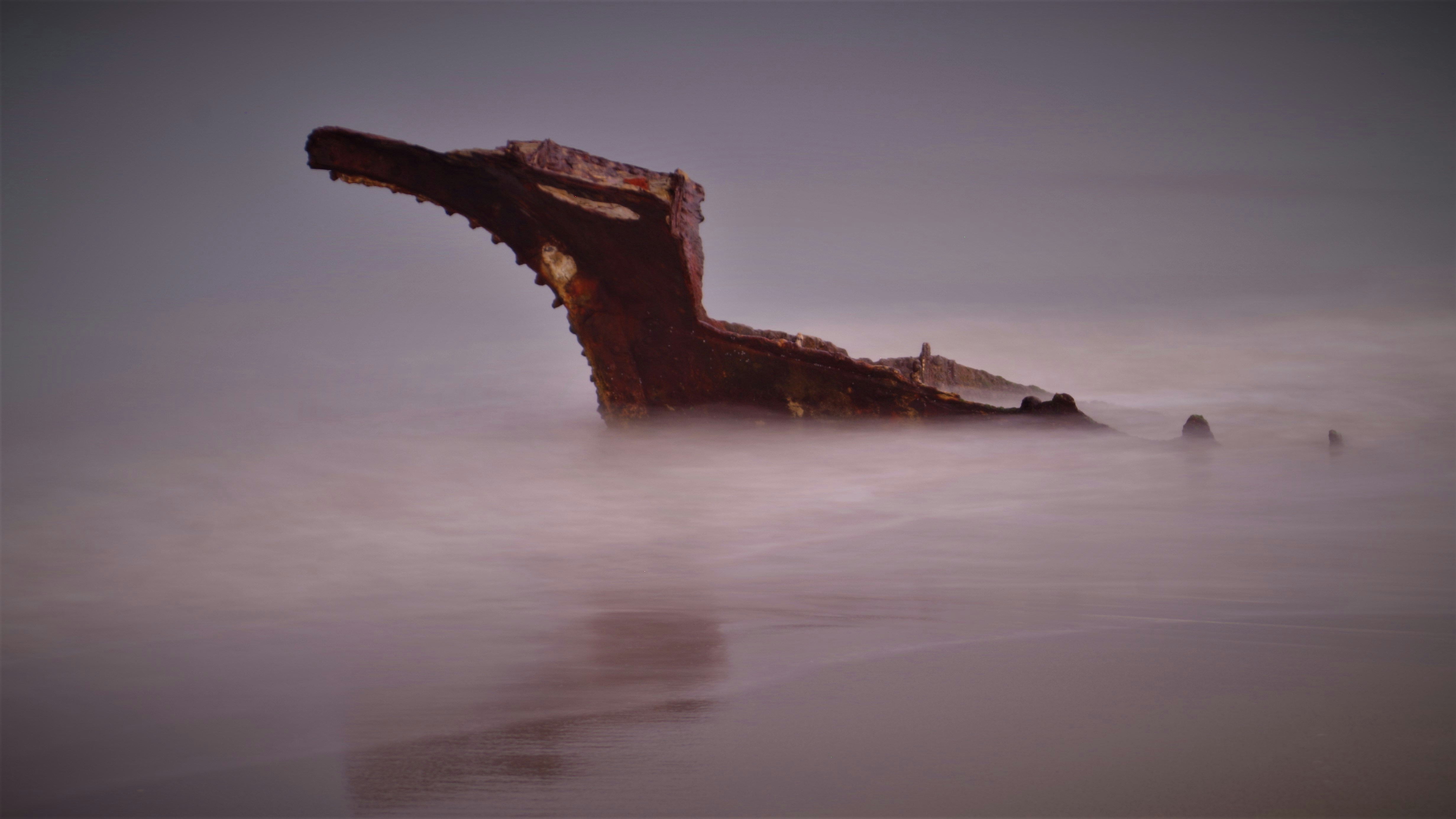 Rusty shipwreck partially submerged in calm waters, surrounded by soft, misty waves.