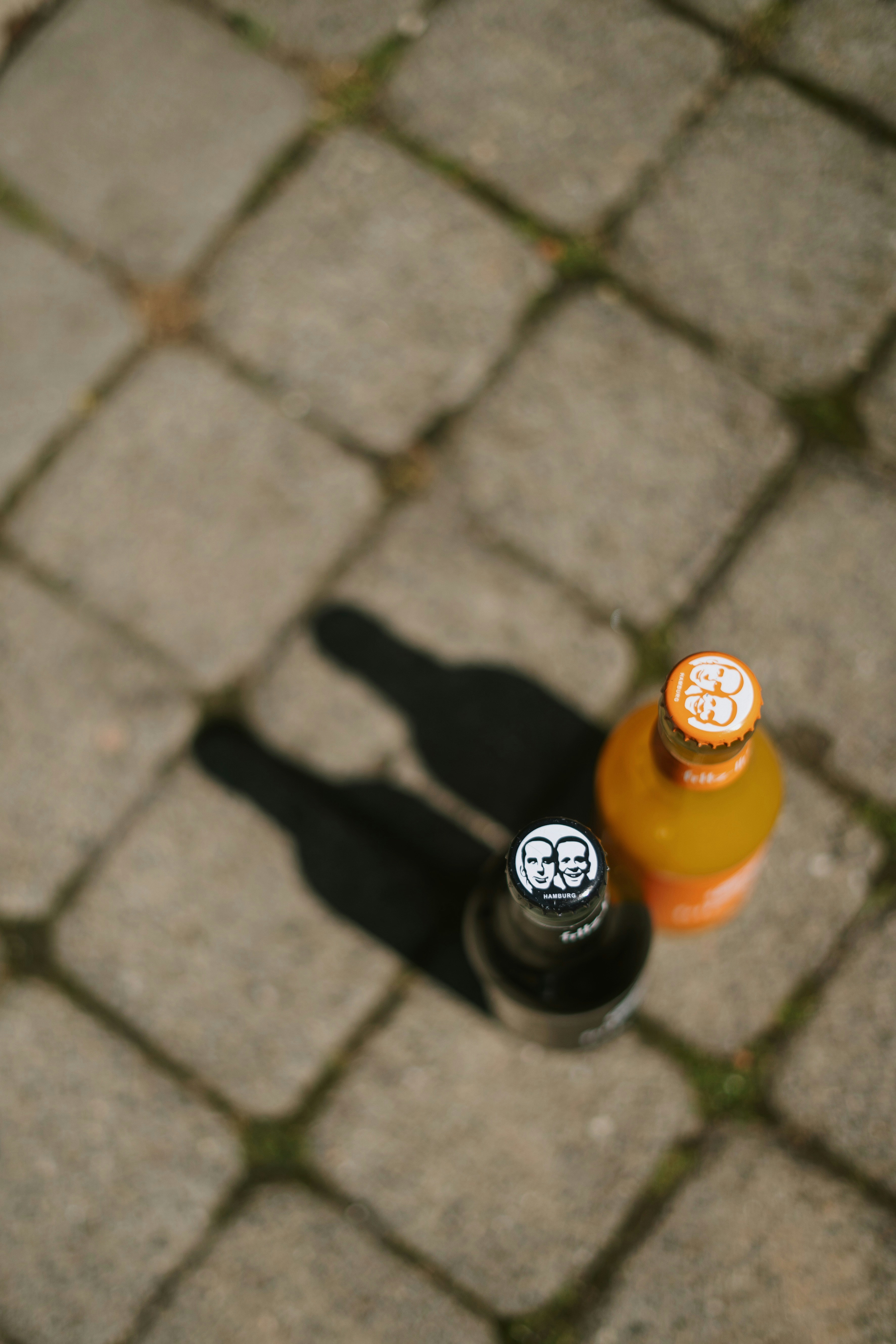 Two beverage bottles cast elongated shadows on a textured cobblestone surface, showcasing distinct caps and colors.