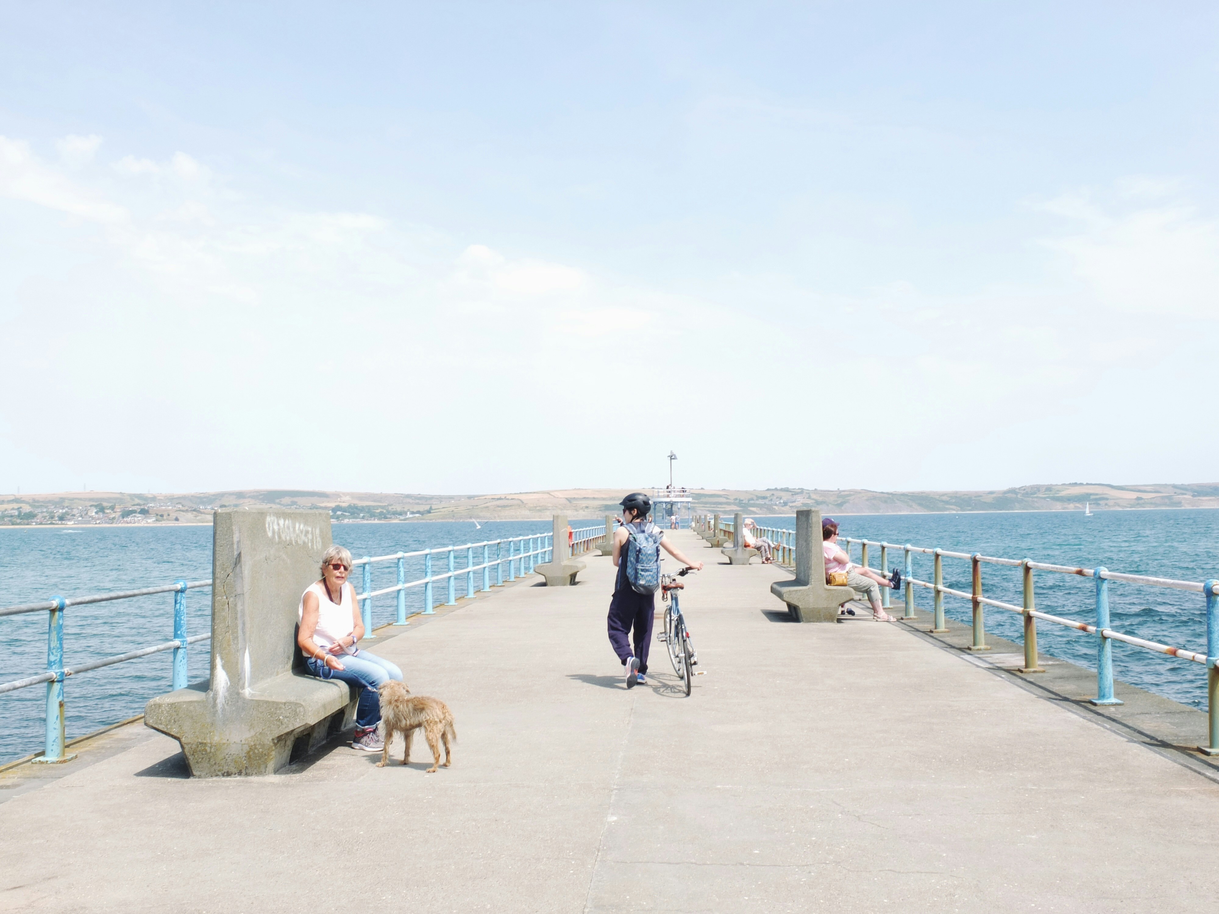 People enjoying a leisurely walk on a sunlit pier overlooking a calm sea.