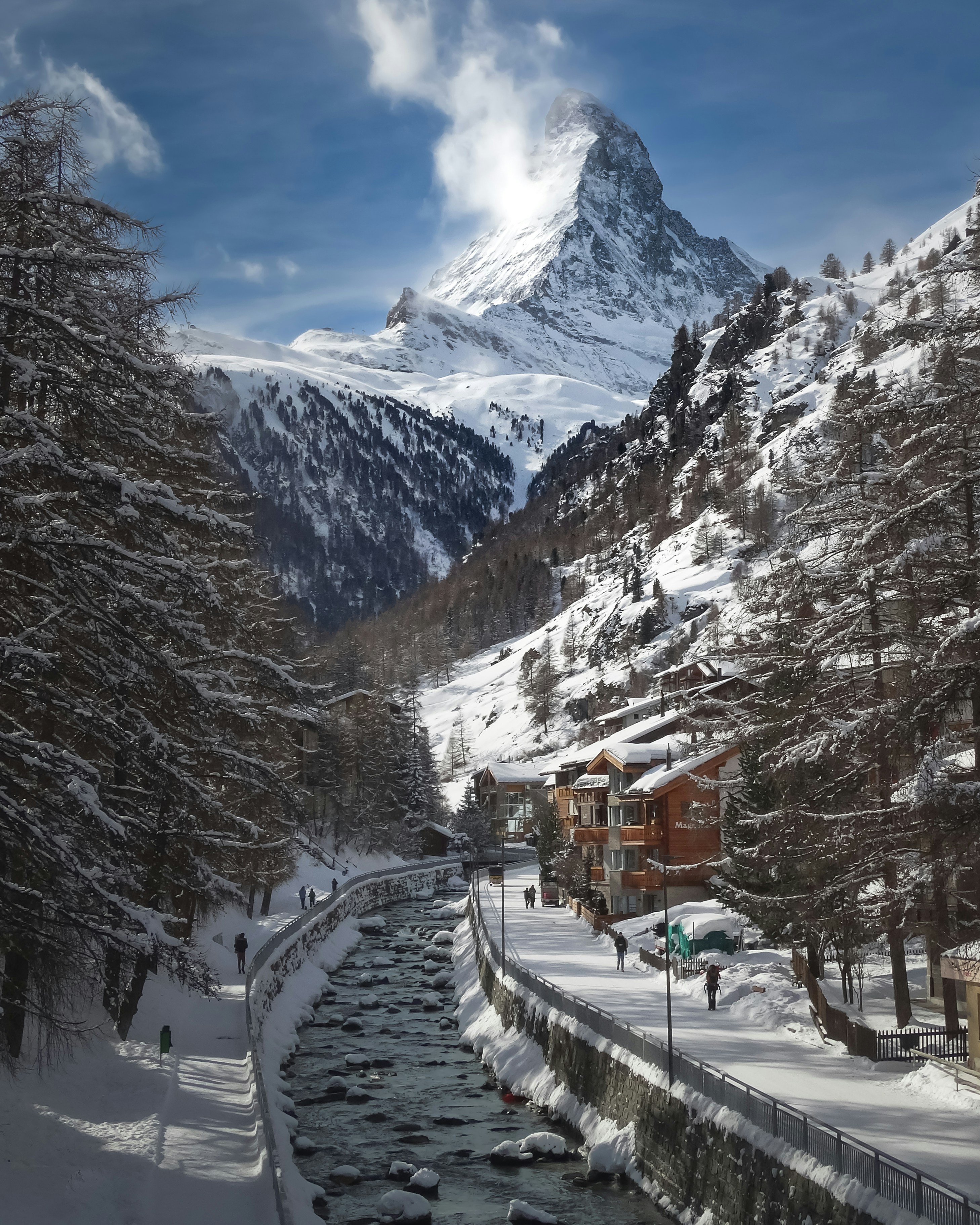 Winter photograph of a snowbound alpine village along a frost-covered river, with a jagged peak looming above the valley.