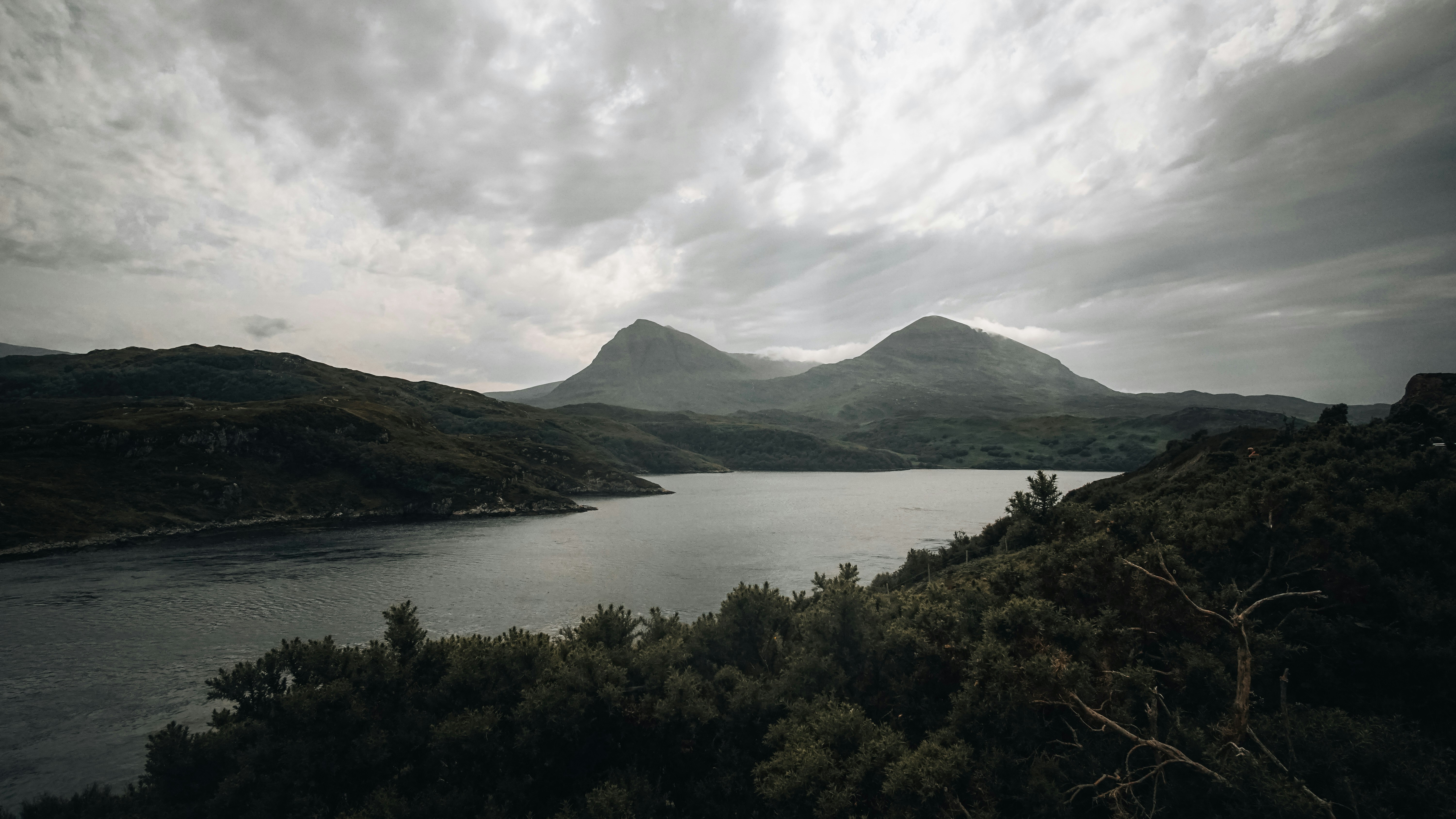 Majestic mountains rise above a tranquil lake, framed by lush greenery under a cloudy sky.