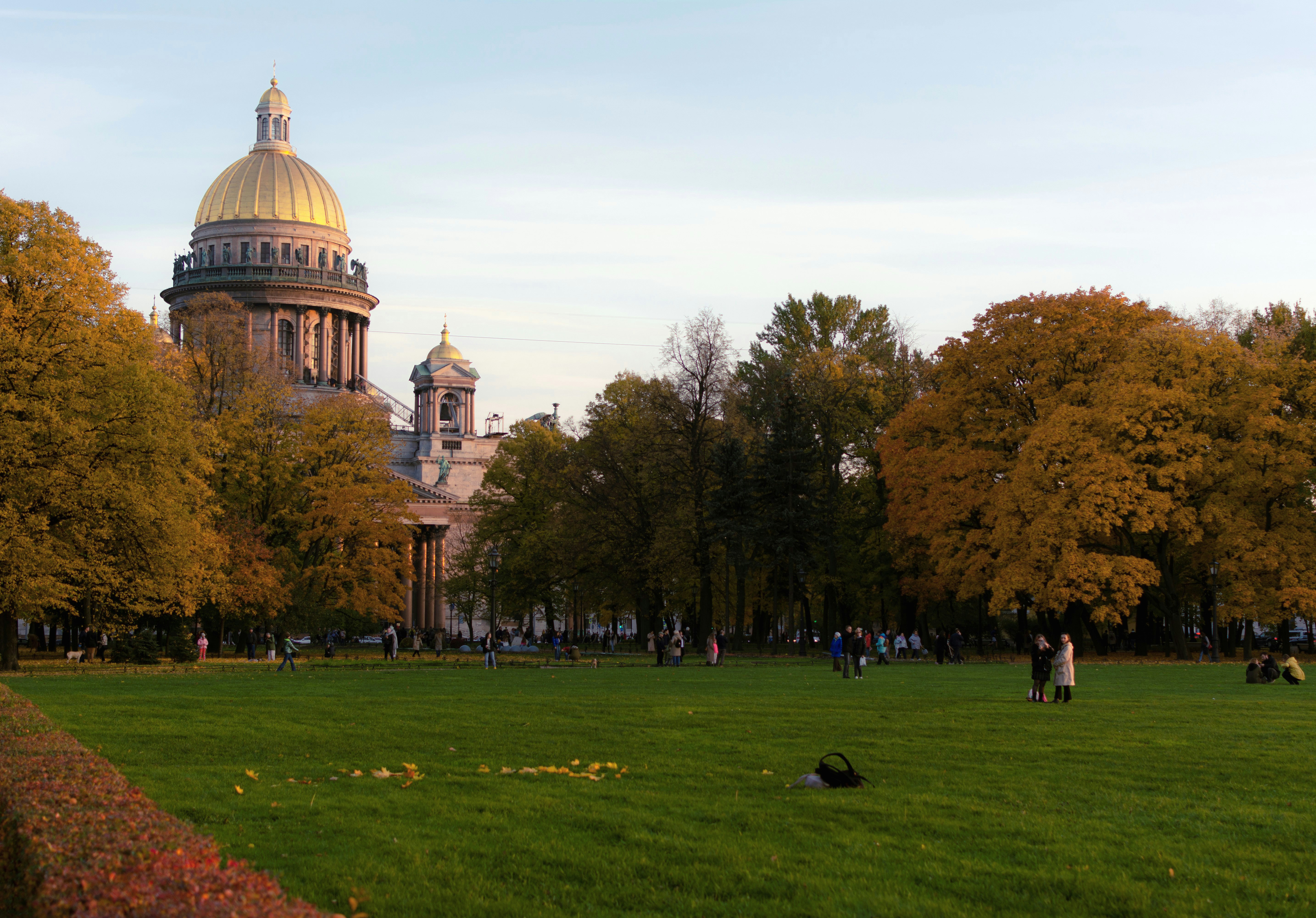 Golden domes of a grand cathedral rise above a vibrant park filled with autumn foliage and leisurely visitors.